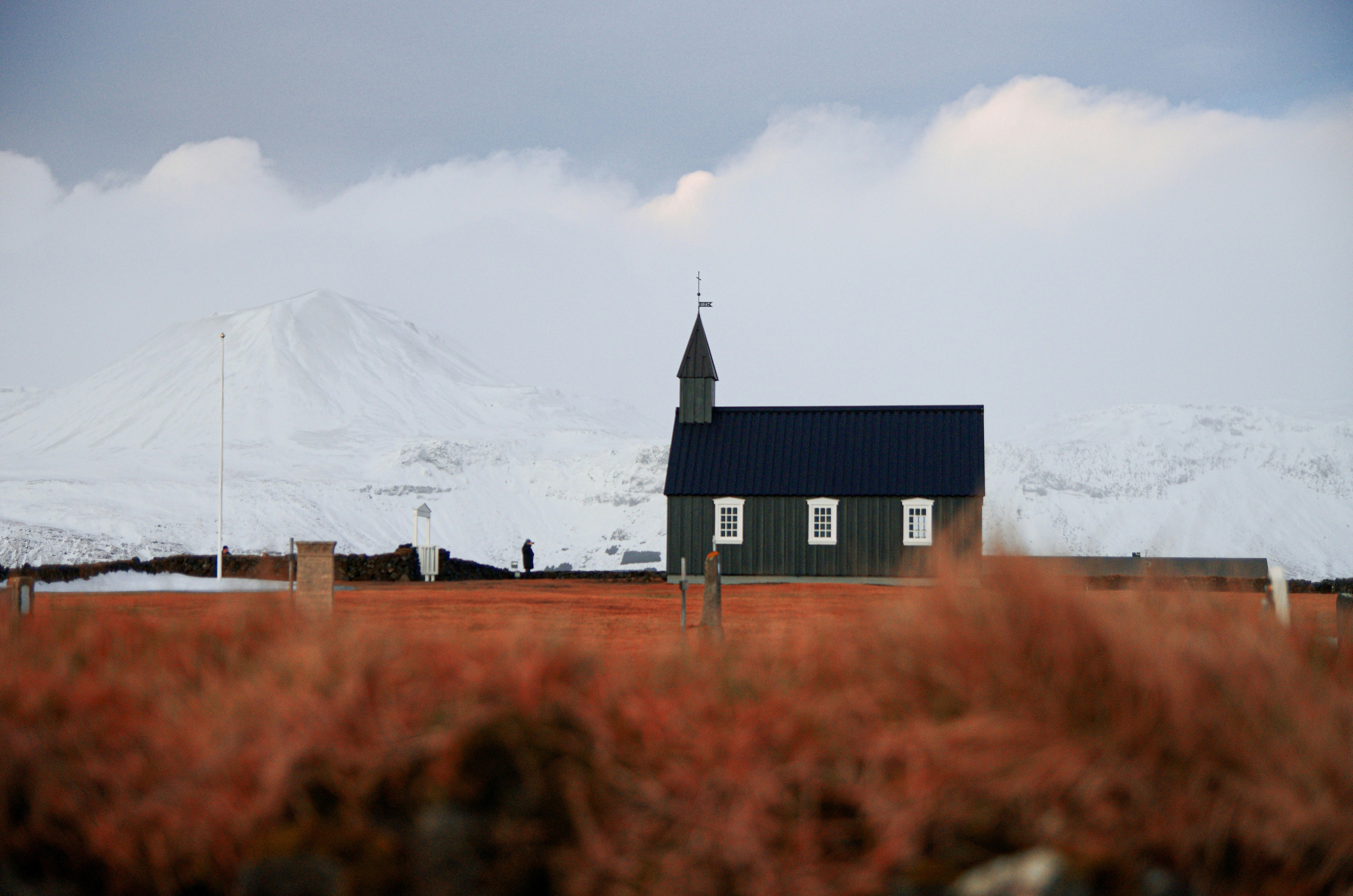 A quaint green church stands against a backdrop of majestic snow-capped mountains, enveloped in soft clouds. The scene conveys a sense of tranquility and isolation.