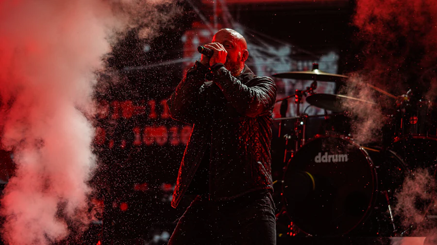 A vocalist sings into a microphone under intense red lighting, with smoke clouds billowing around the stage and a drum kit behind them.