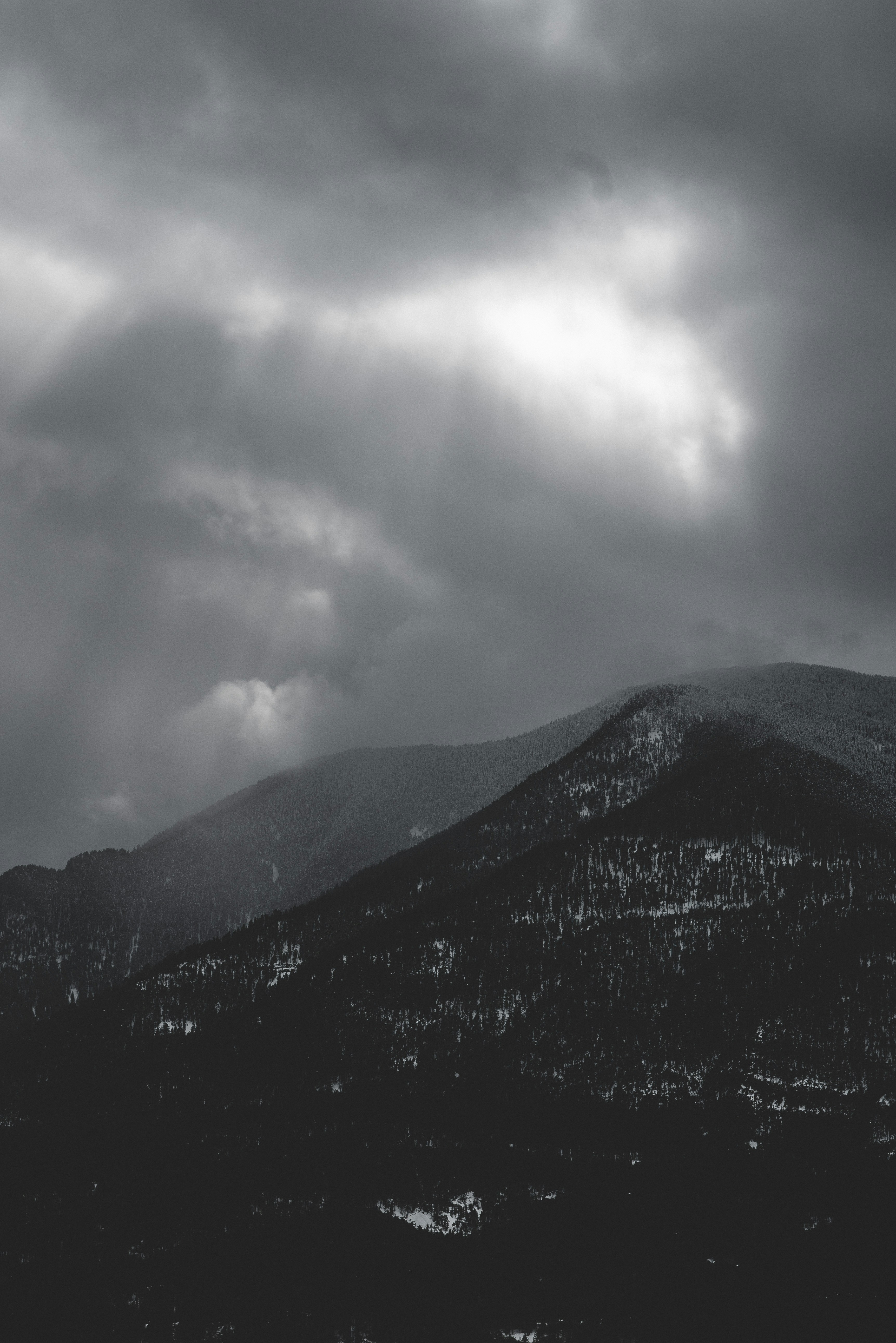 Dark, brooding mountains under a tumultuous sky, with rays of light piercing through the clouds. Snow blankets the terrain, enhancing the scene's stark beauty.