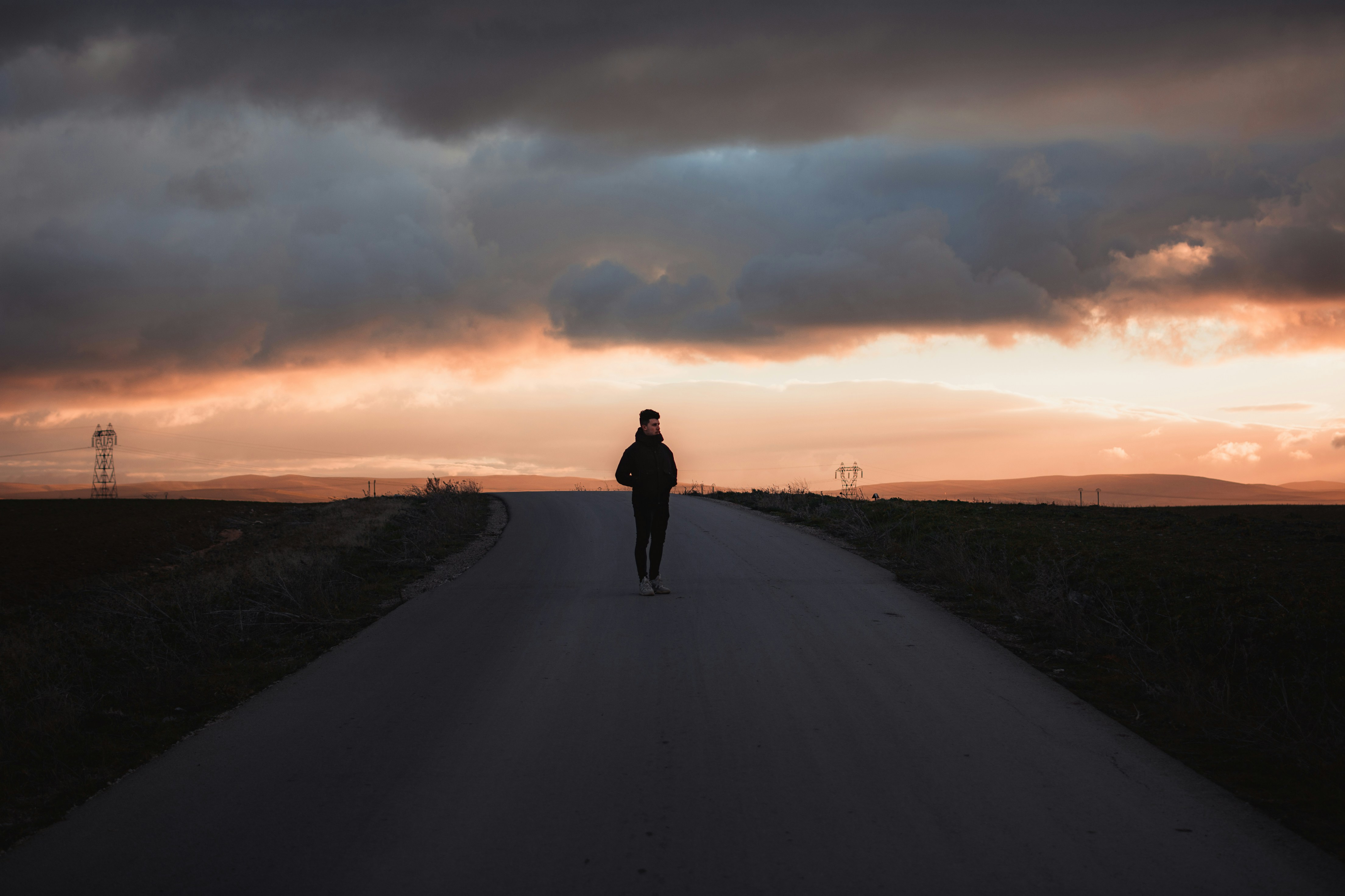 Silhouette of a person walking along a deserted road beneath dramatic clouds at sunset.