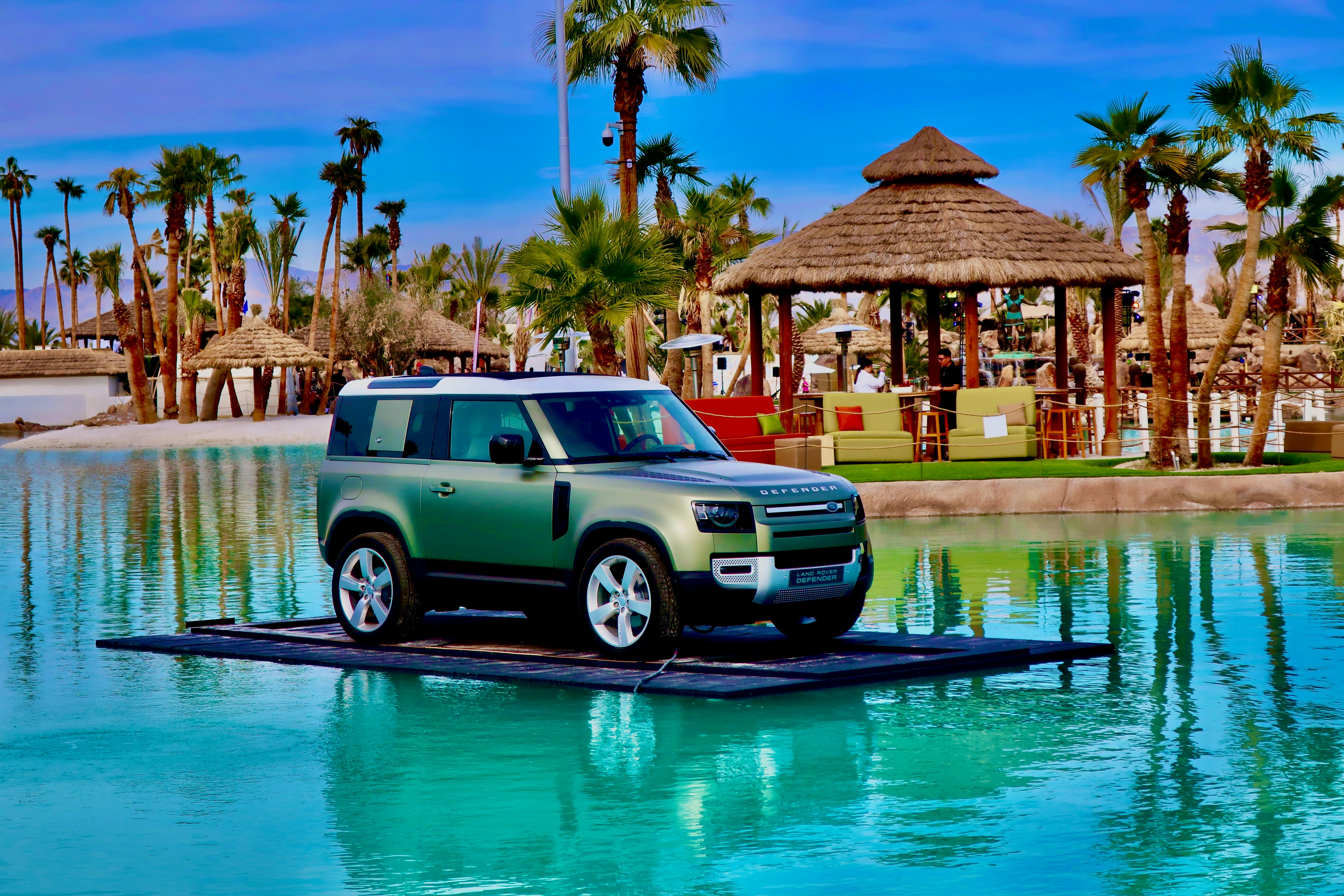 Green SUV on a platform surrounded by clear turquoise water with palm trees and a thatched pavilion in the background.