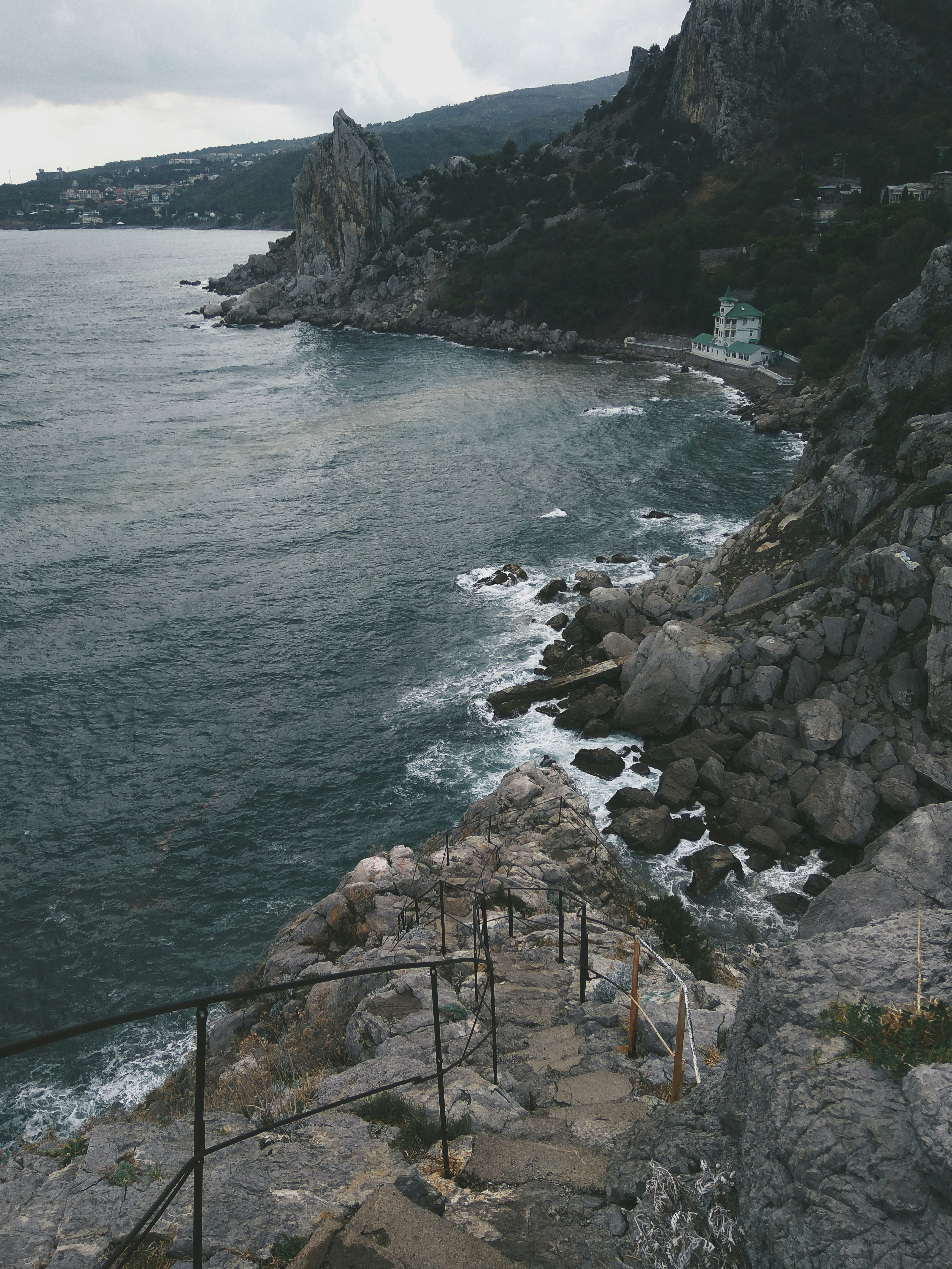 Winding stone steps lead down to a rugged coastline where waves crash against rocks, framed by a distant green building and dramatic cliffs.