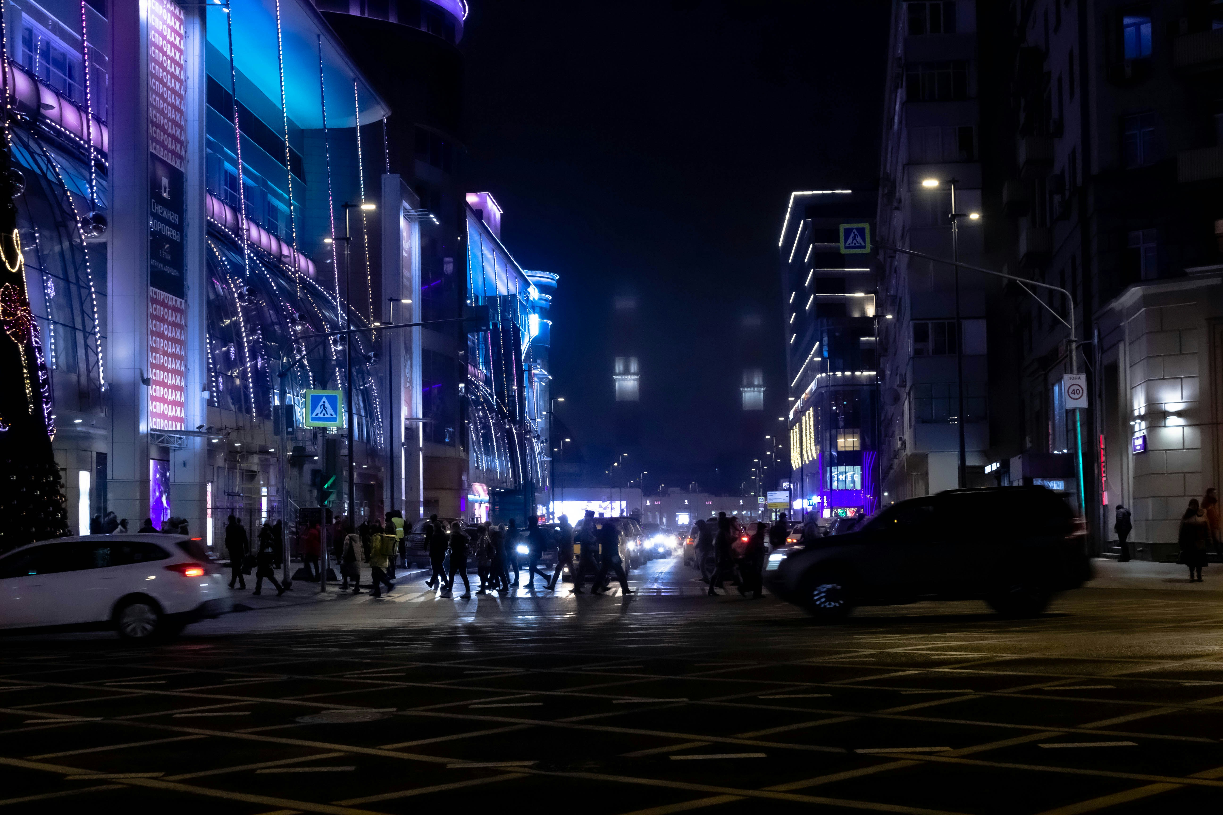 Vibrant city street illuminated by colorful lights, showcasing pedestrians crossing amidst modern architecture at night.