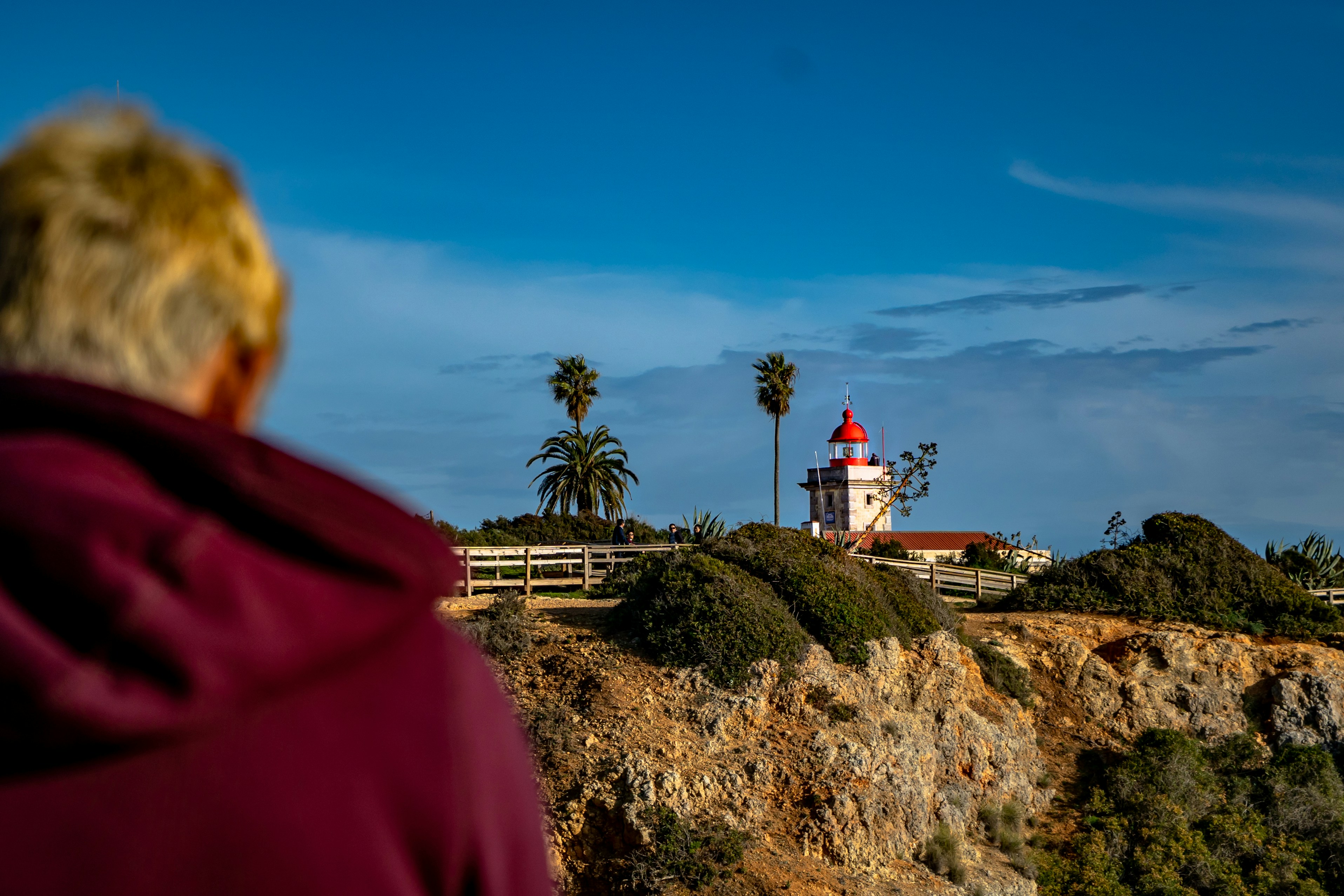 Person in red hoodie gazing at a distant lighthouse set against a clear blue sky.