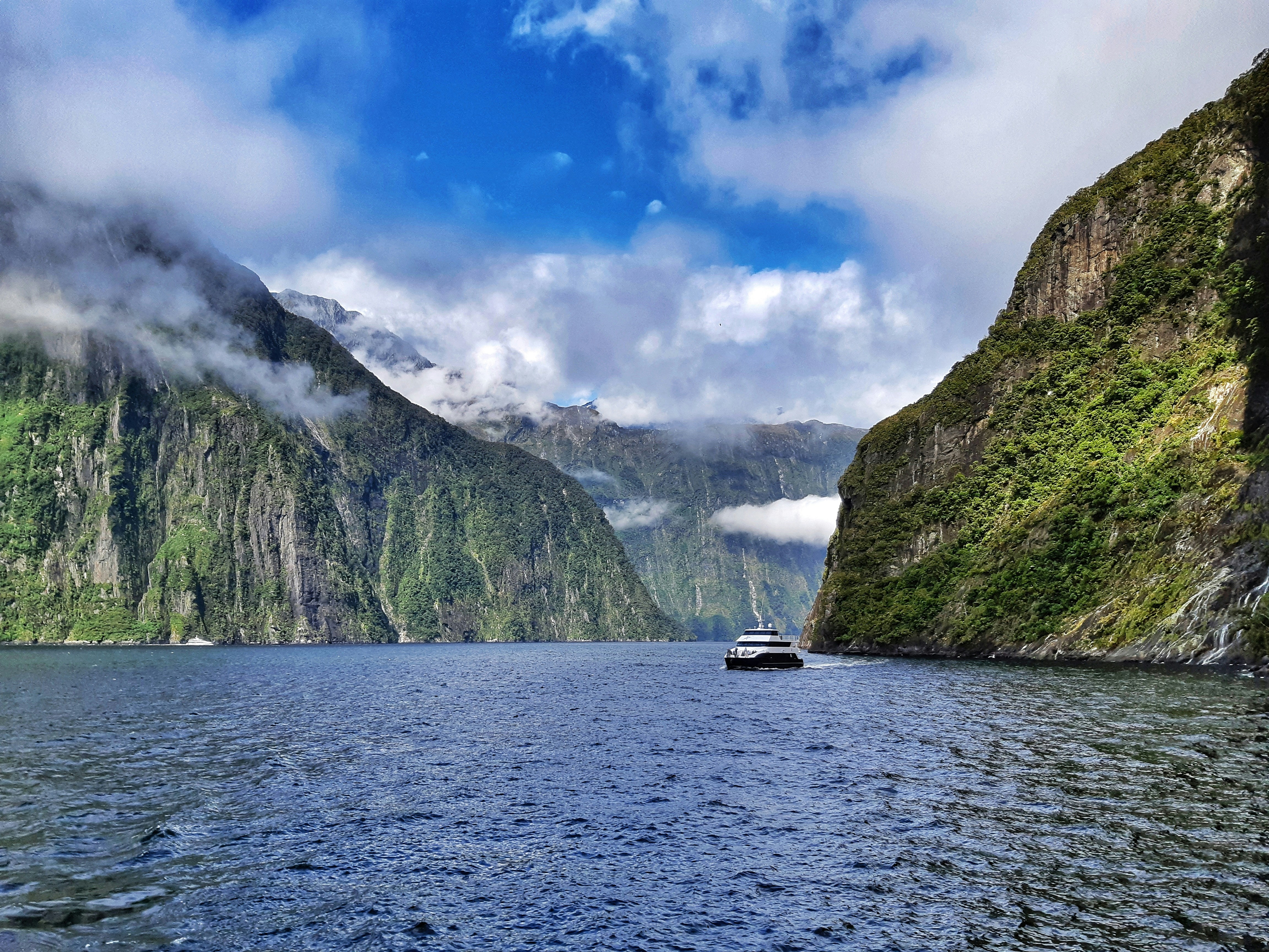 A boat glides through a tranquil fjord, surrounded by towering cliffs and lush greenery under a partly cloudy sky.