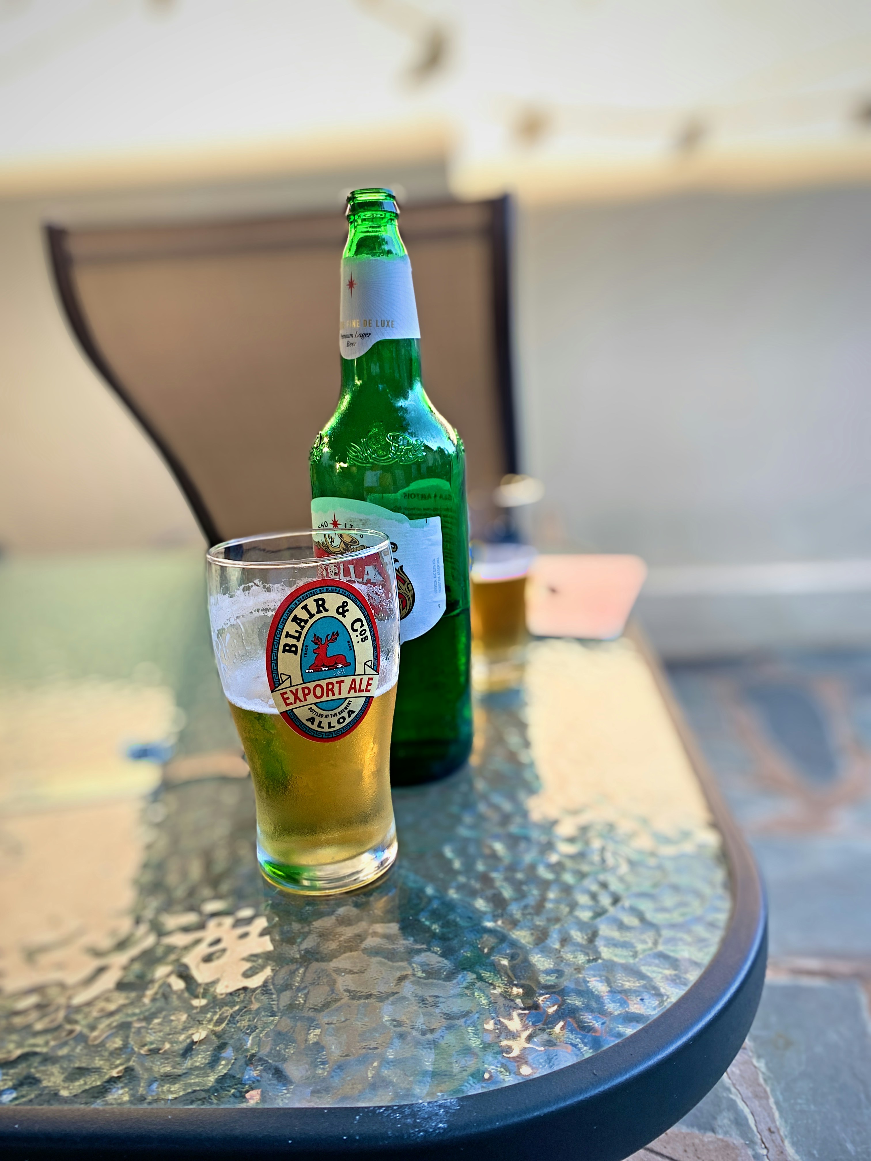 A glass of beer with frothy top sits beside a green bottle on a textured glass table, evoking a relaxed outdoor atmosphere.