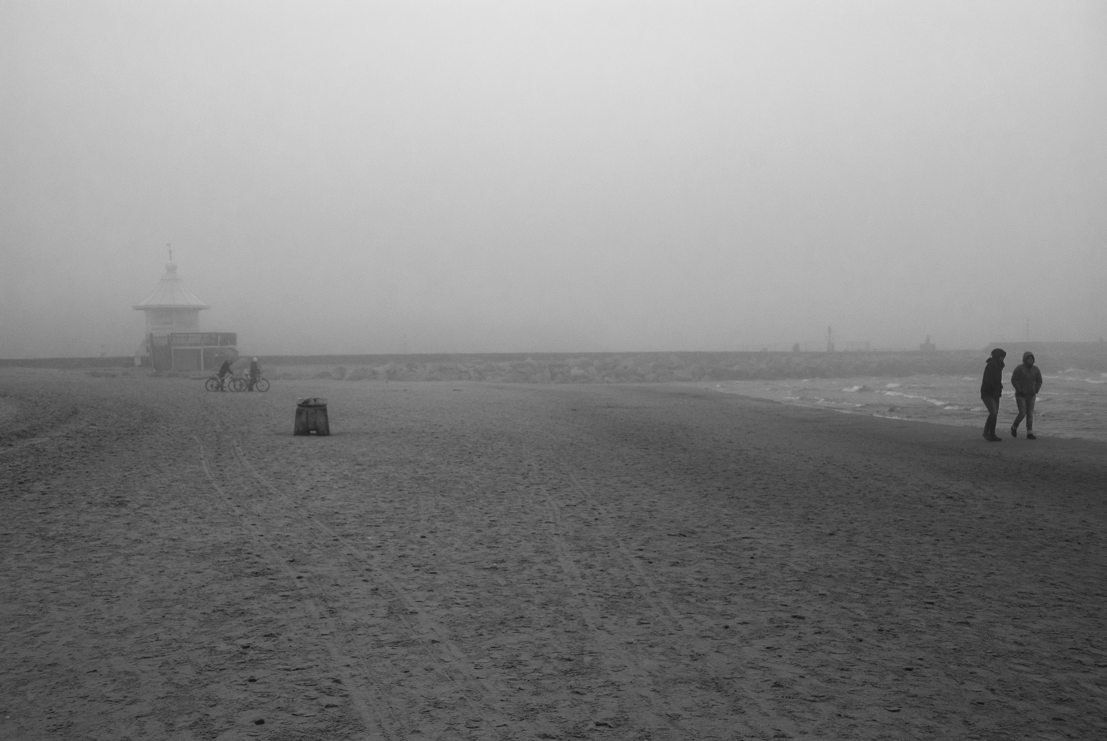 Grayscale scene of a beach with a distant lifeguard tower and two figures walking through fog.