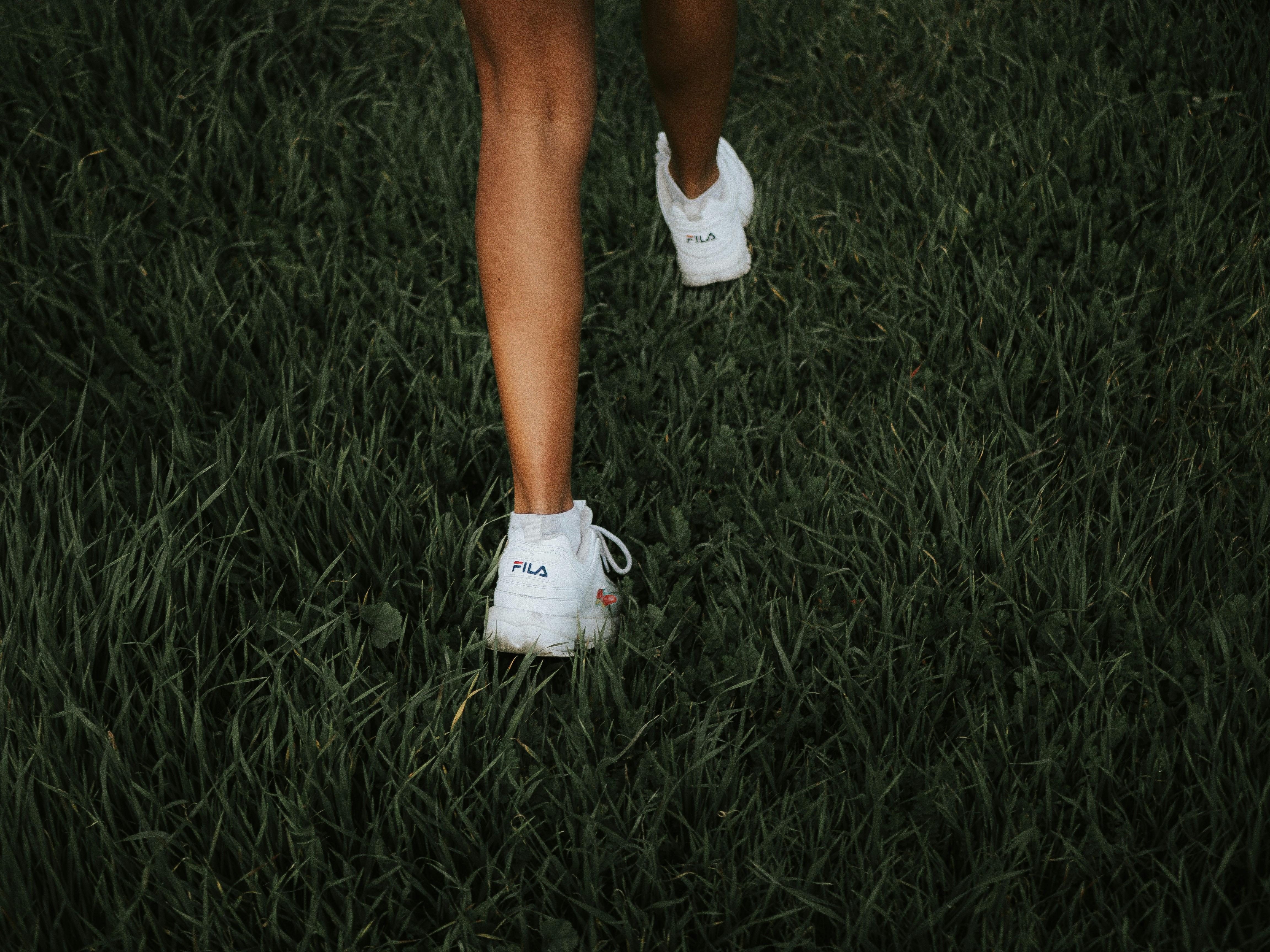 A person walks through lush green grass, showcasing white FILA sneakers against the vibrant backdrop of nature.