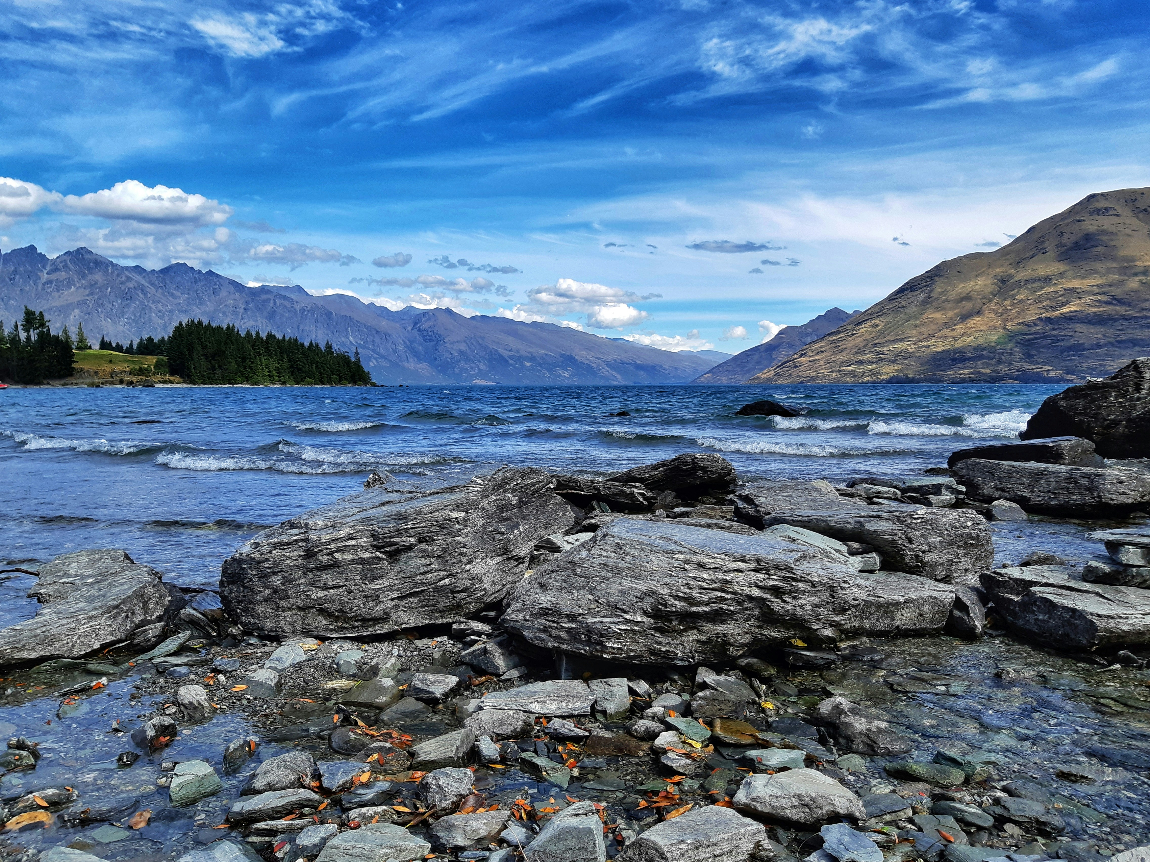 Rocky shoreline of Lake Wakatipu with distant mountains under a vibrant blue sky.