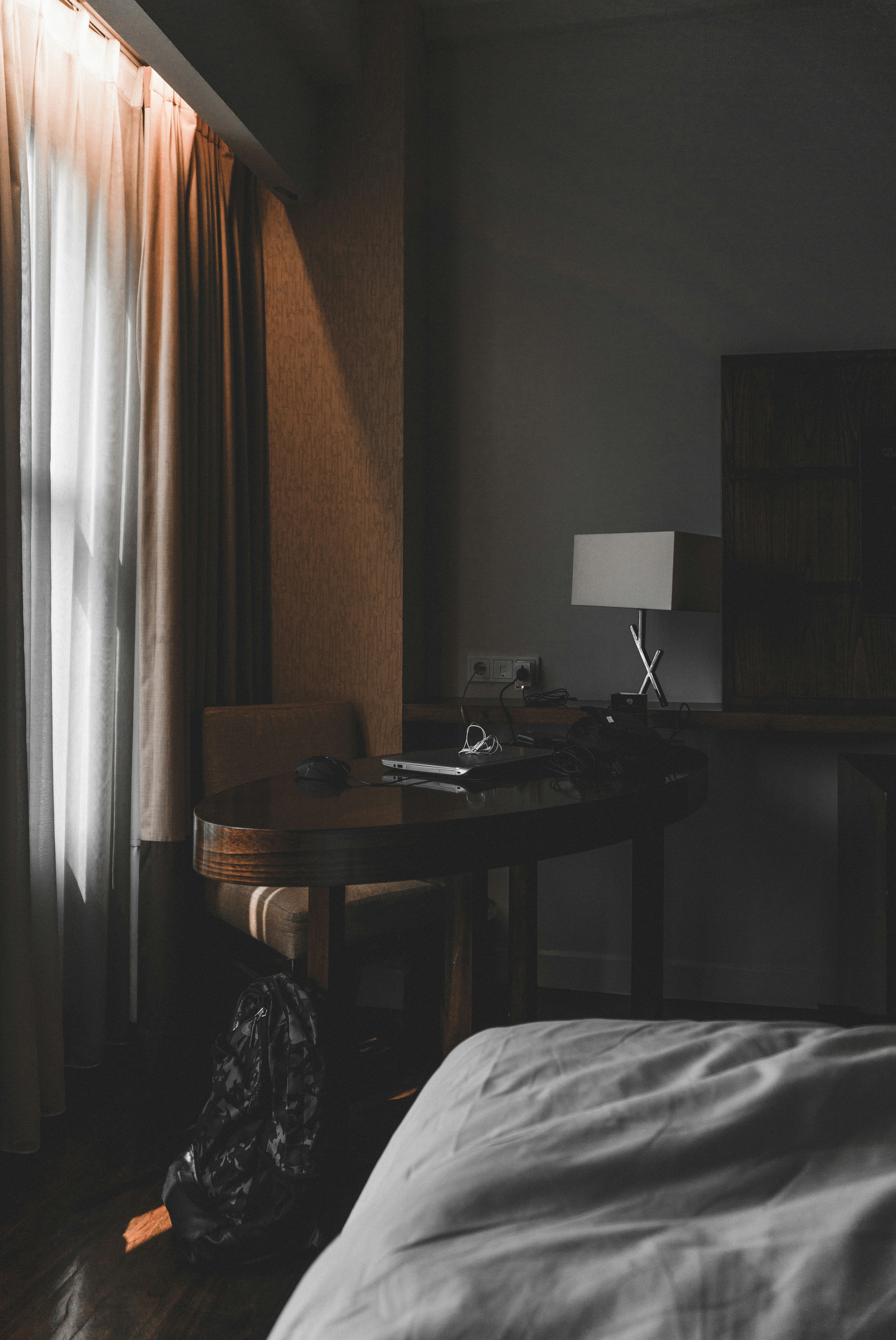 A serene hotel room scene featuring a wooden table with a laptop and a chair, softly illuminated by natural light filtering through the curtains.