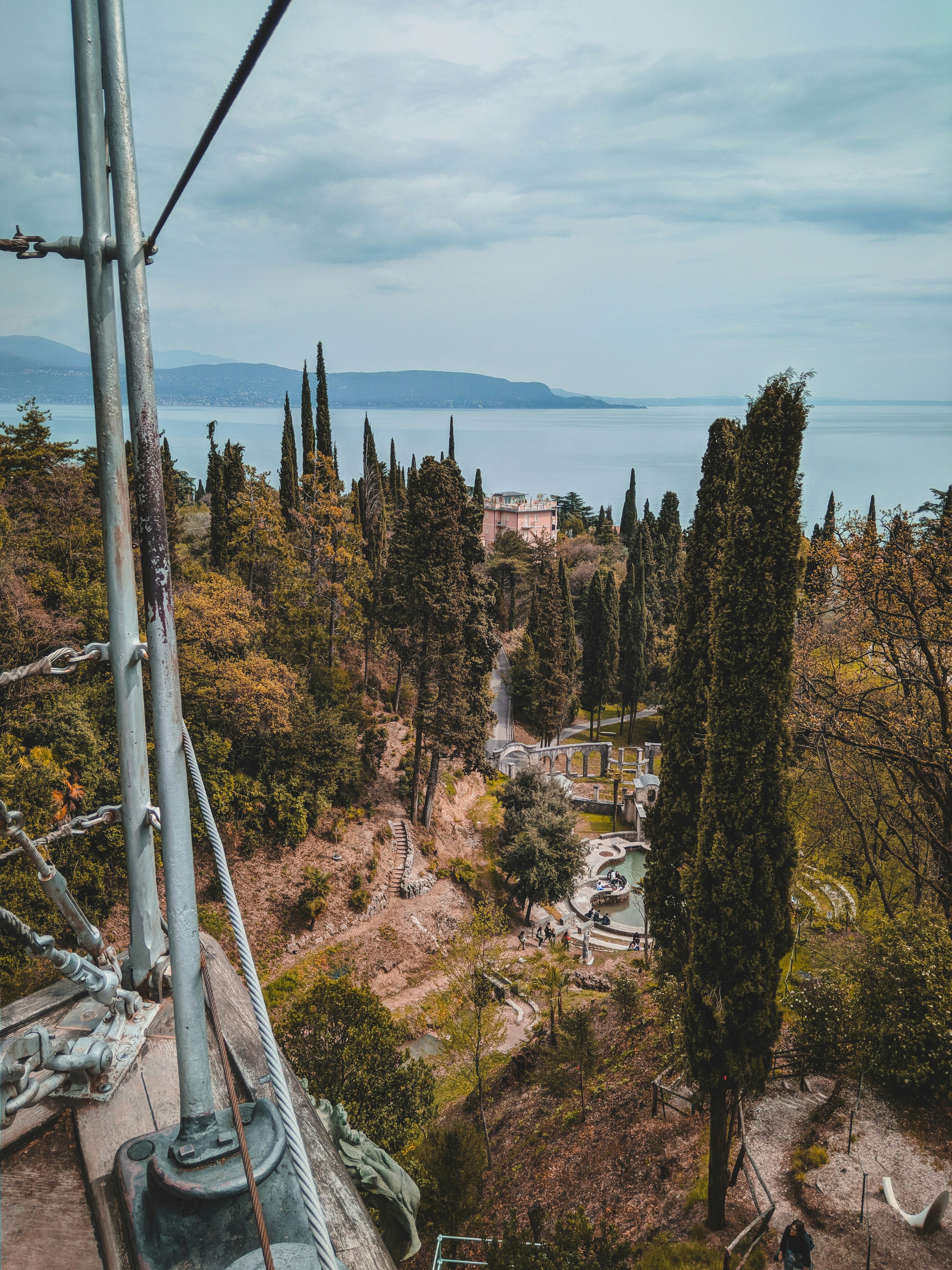 Terraced hillside park with winding paths and tall cypress trees leads toward a calm bay on the horizon. The photo is taken from an elevated vantage near scaffolding, emphasizing the overlook.