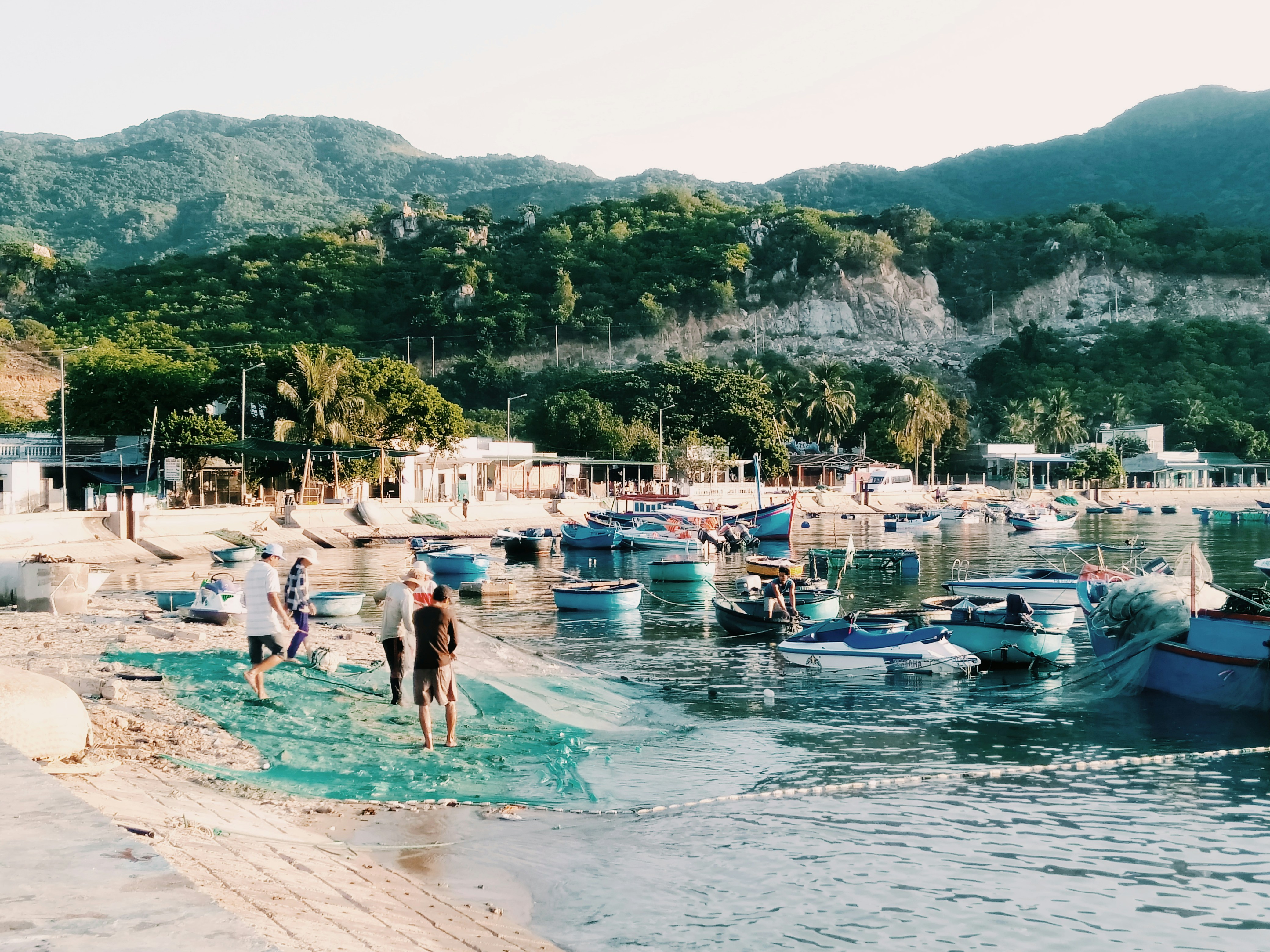 Small marina with colorful boats docked along a calm shoreline. Two people stroll on a green-tinted dock with tree-covered hills rising in the background.