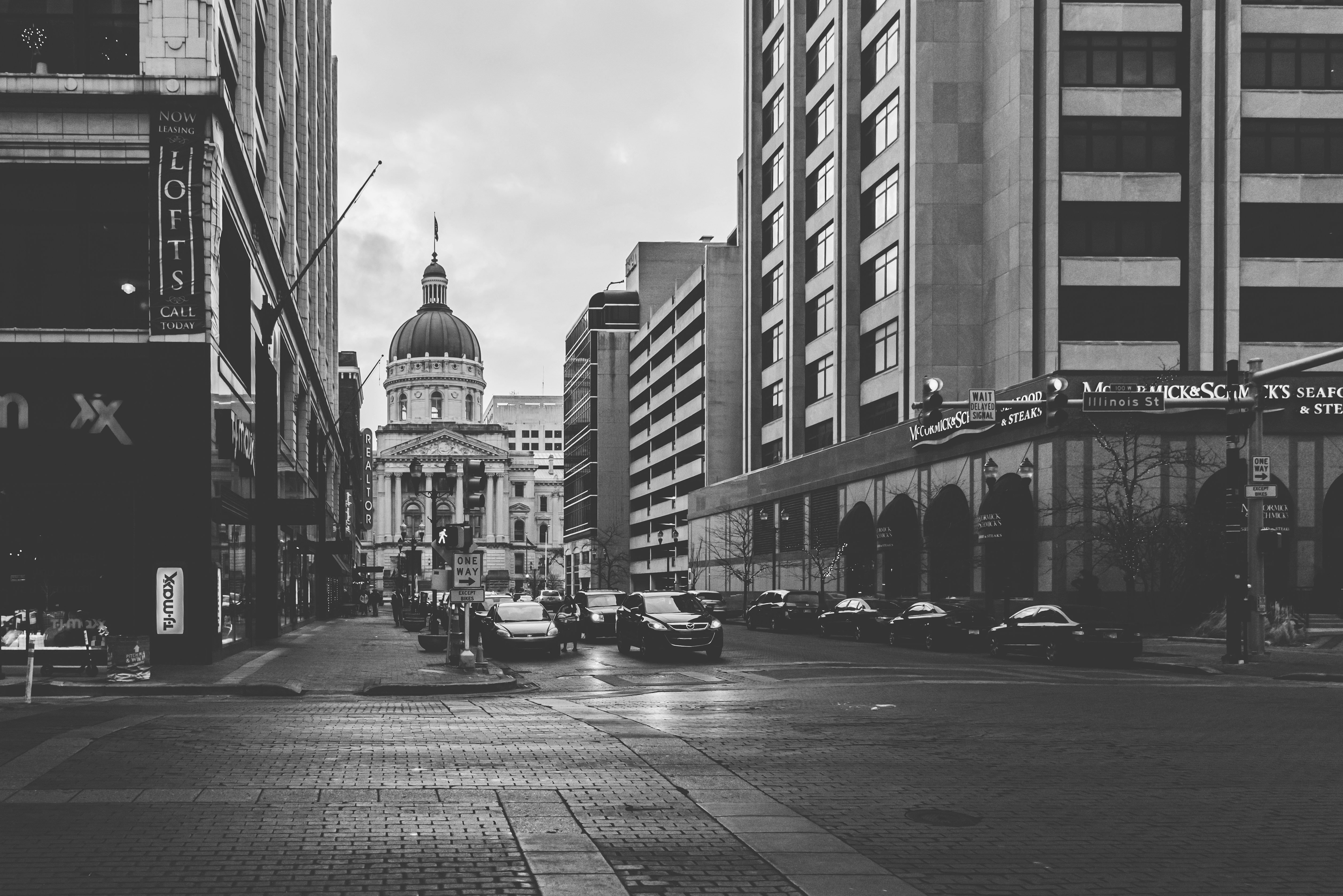 Grayscale scene of cars parked along a city street framed by towering high-rise buildings.