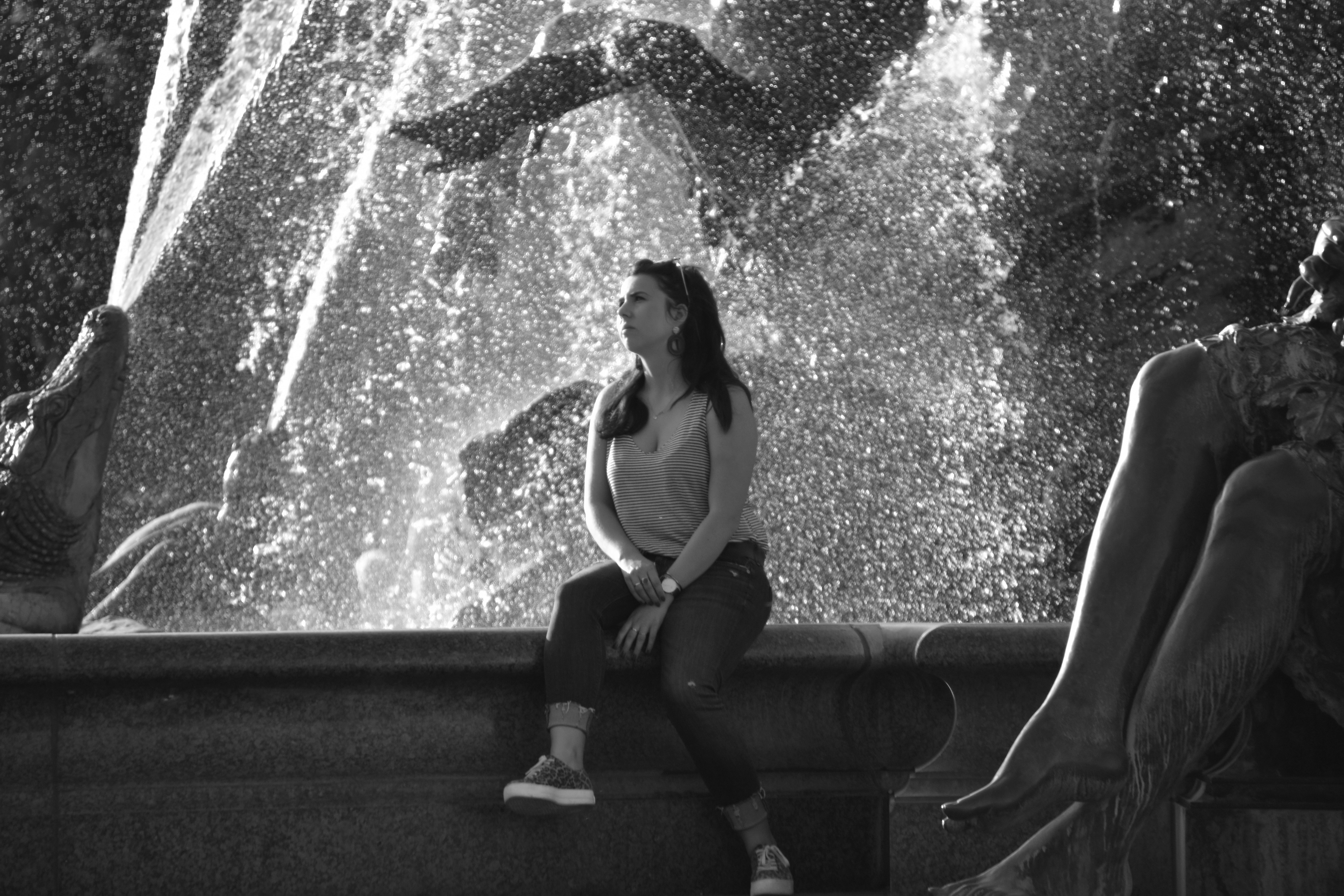 A woman sits pensively by a fountain, surrounded by splashes of water in a monochrome setting.