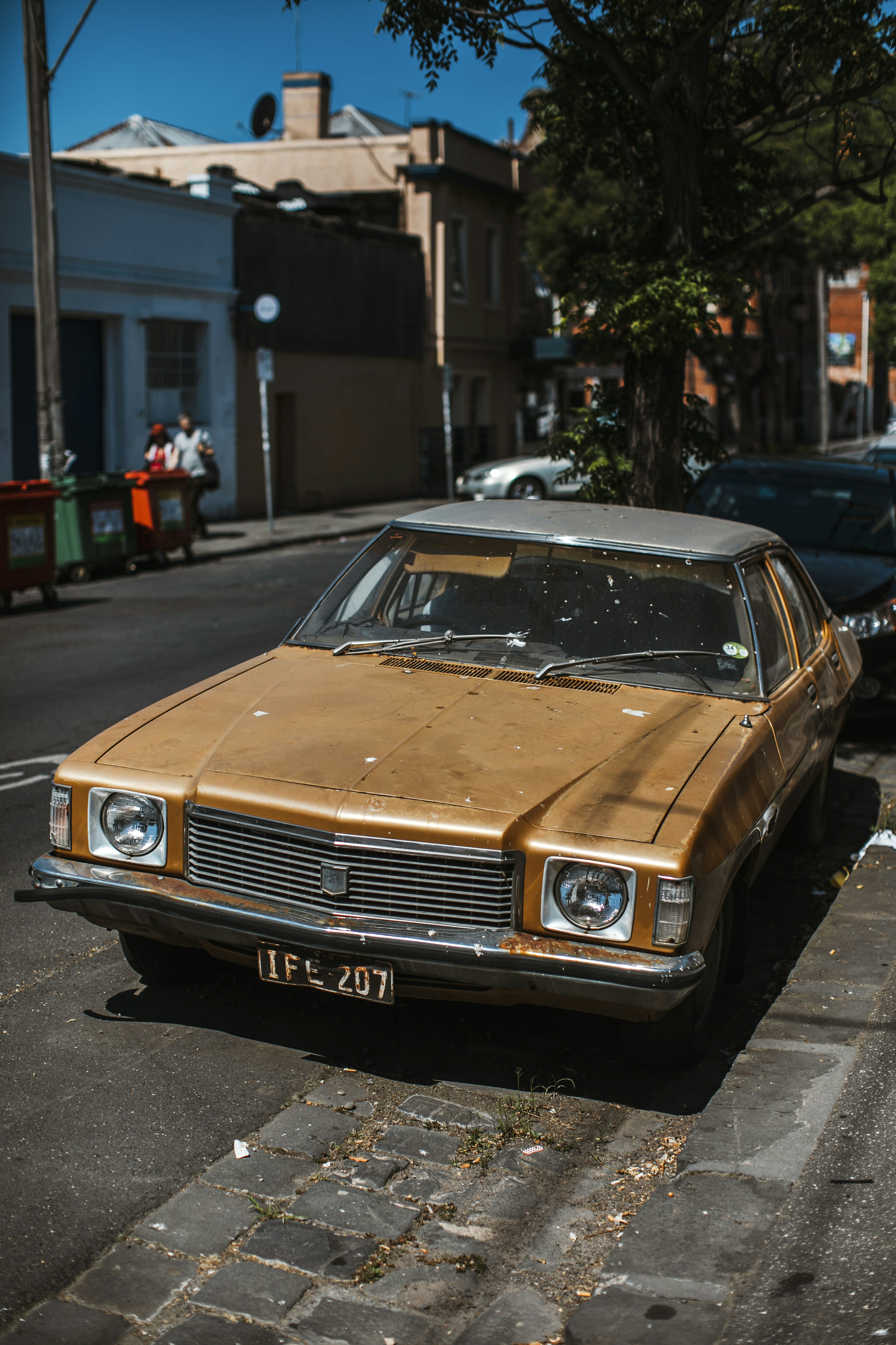 Weathered gold car parked on a sunlit street, showcasing its aged charm amidst urban surroundings.