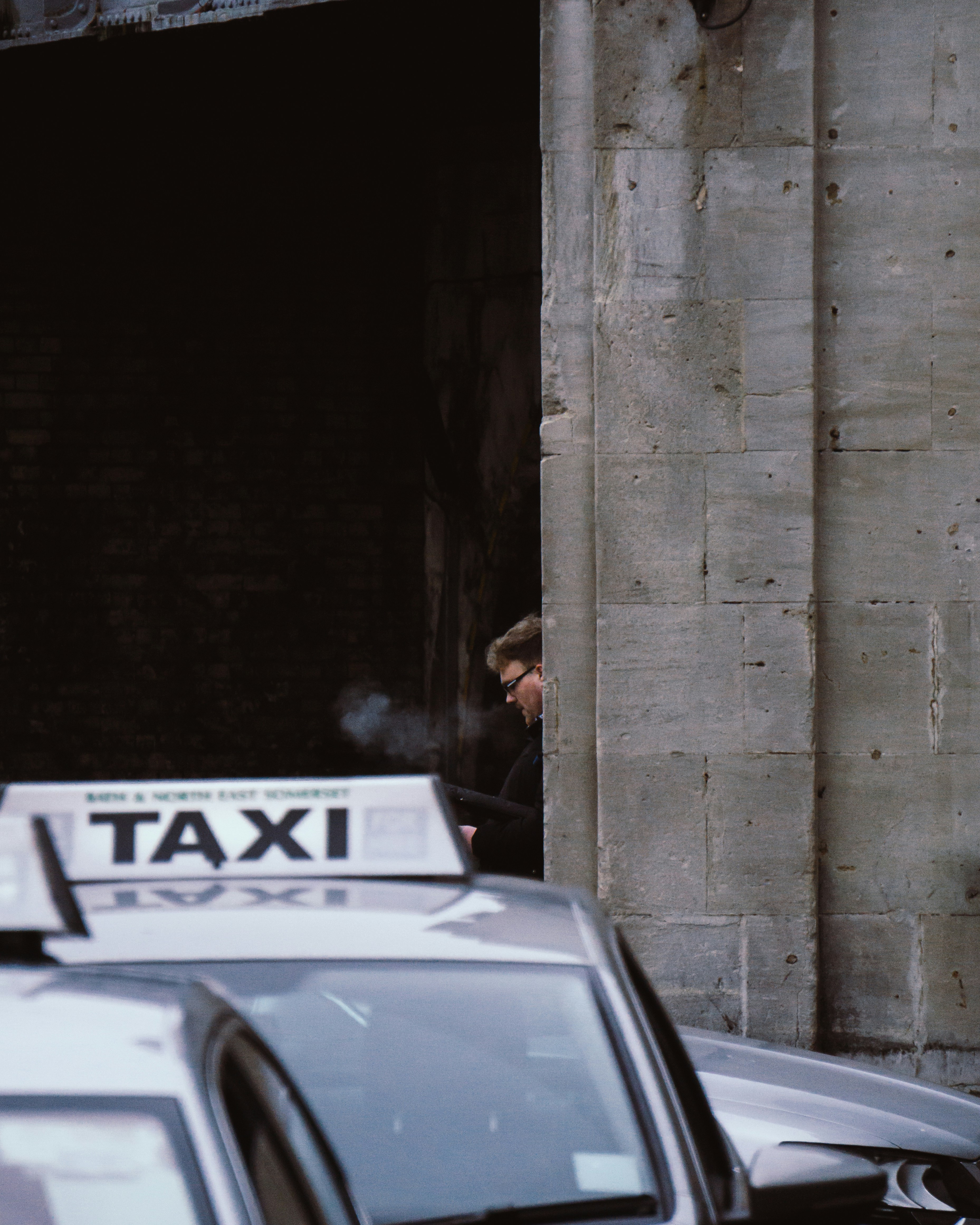 A man stands in the shadows near a stone wall, exhaling smoke, while taxis wait in the foreground. The scene captures a fleeting moment of urban life.