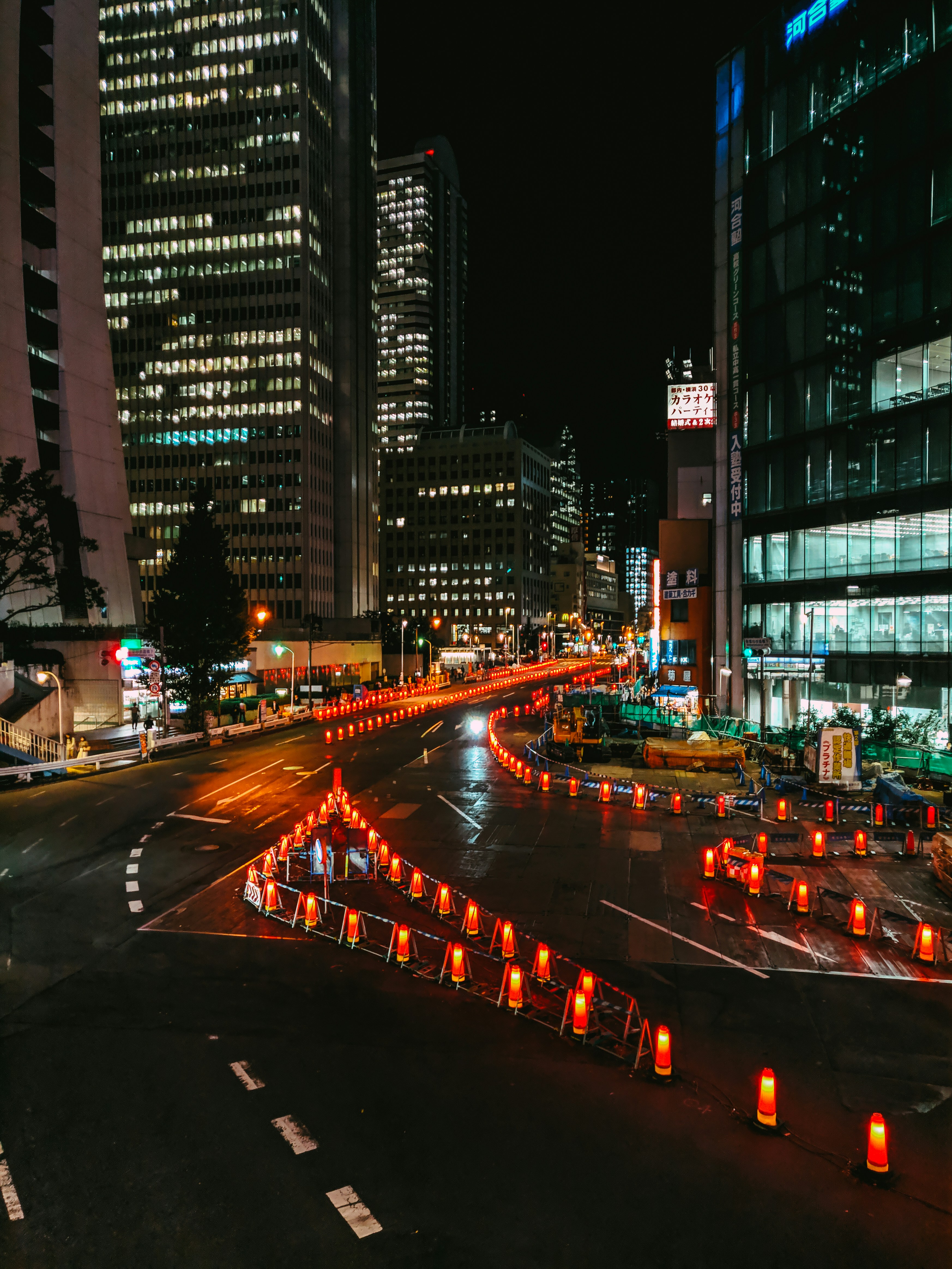 Illuminated traffic cones line a winding road in a bustling city at night, framed by towering skyscrapers. The scene captures the vibrant energy of urban life.