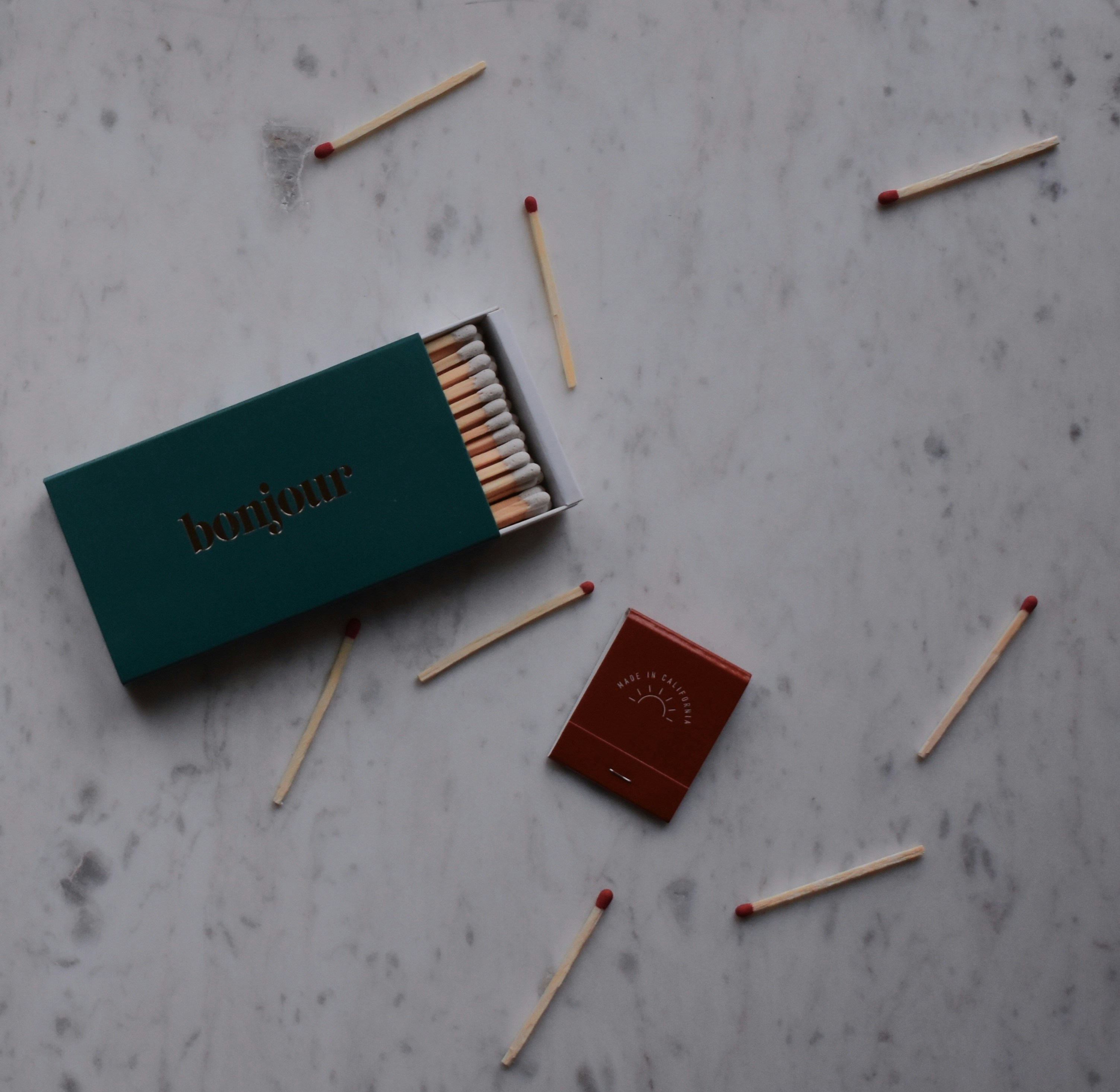A teal matchbox labeled 'bonjour' surrounded by scattered matchsticks on a marble surface.