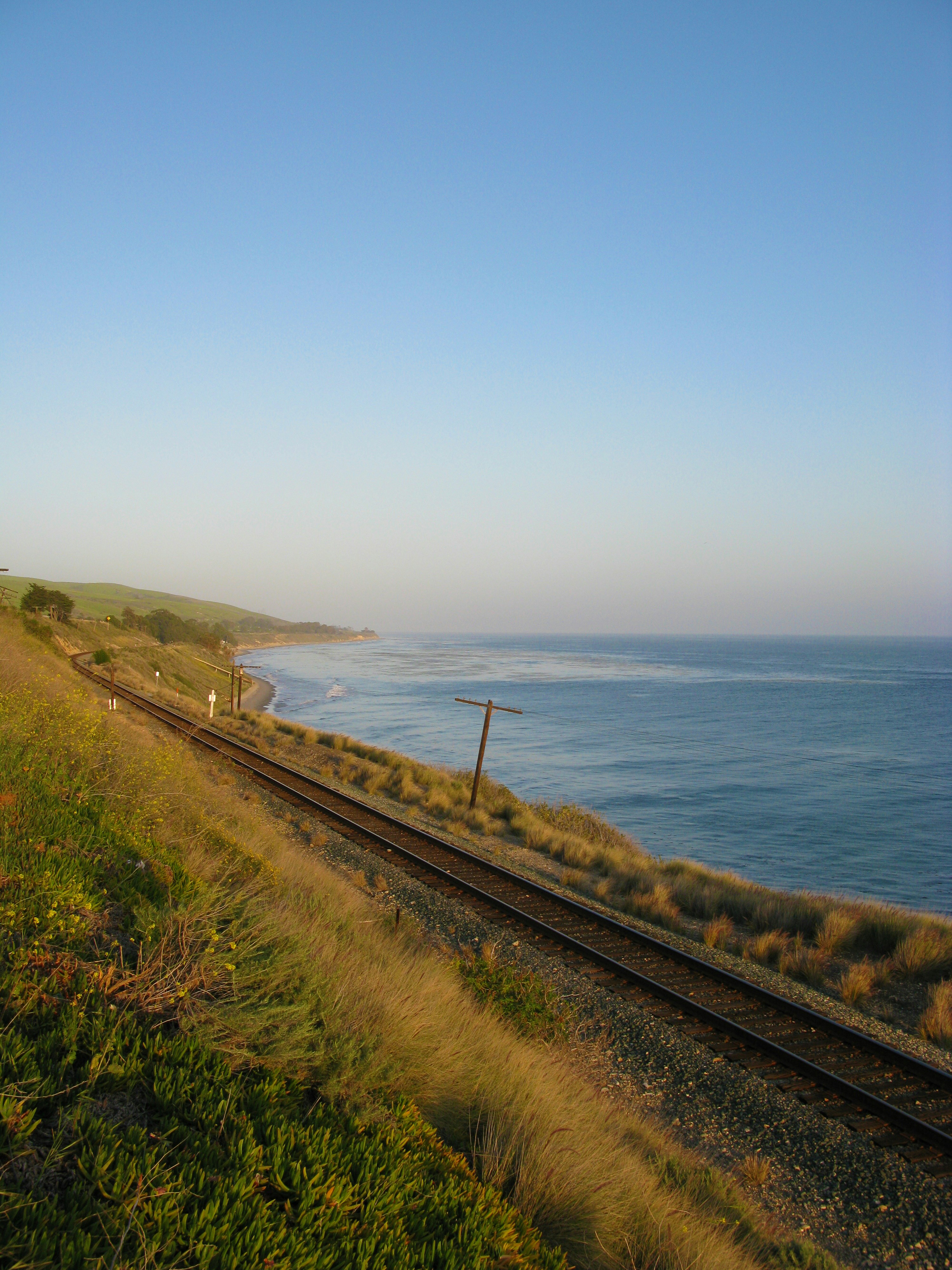 Winding railway tracks alongside a serene coastline under a clear sky, with gentle waves lapping at the shore.
