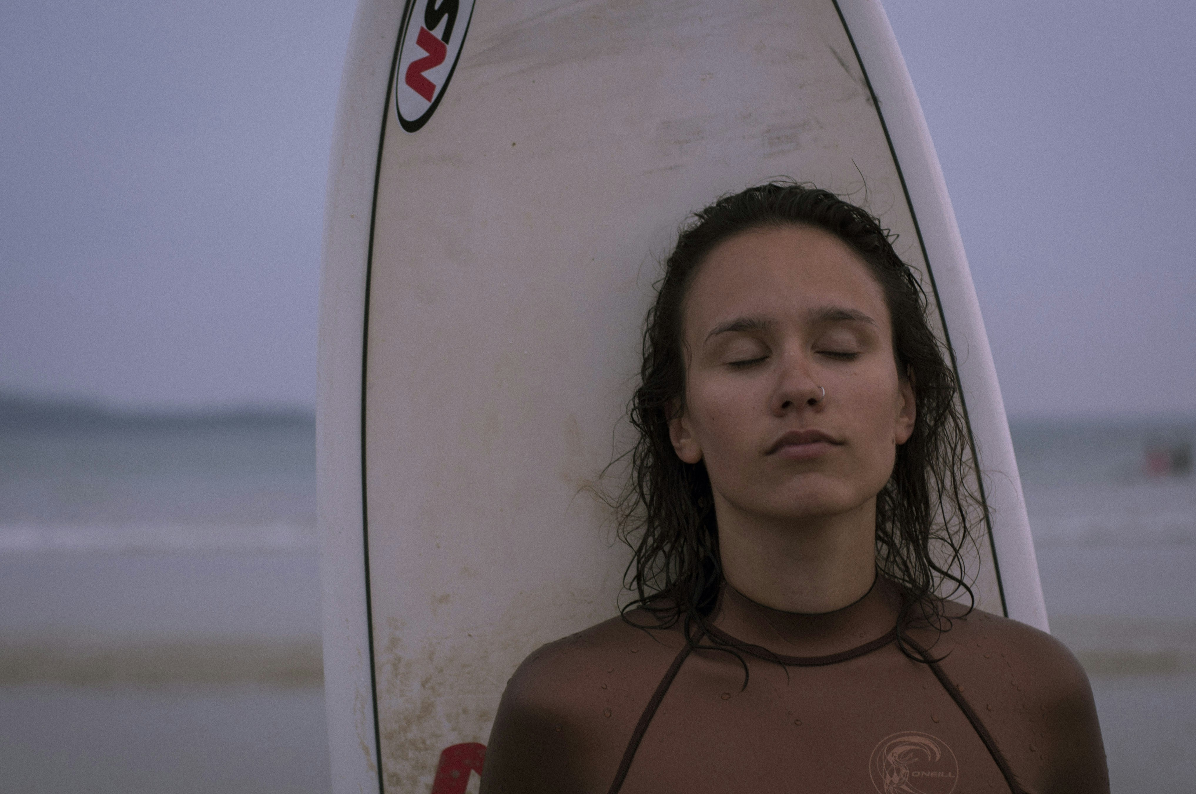 Surfer with closed eyes resting against a surfboard on a tranquil beach at dusk.