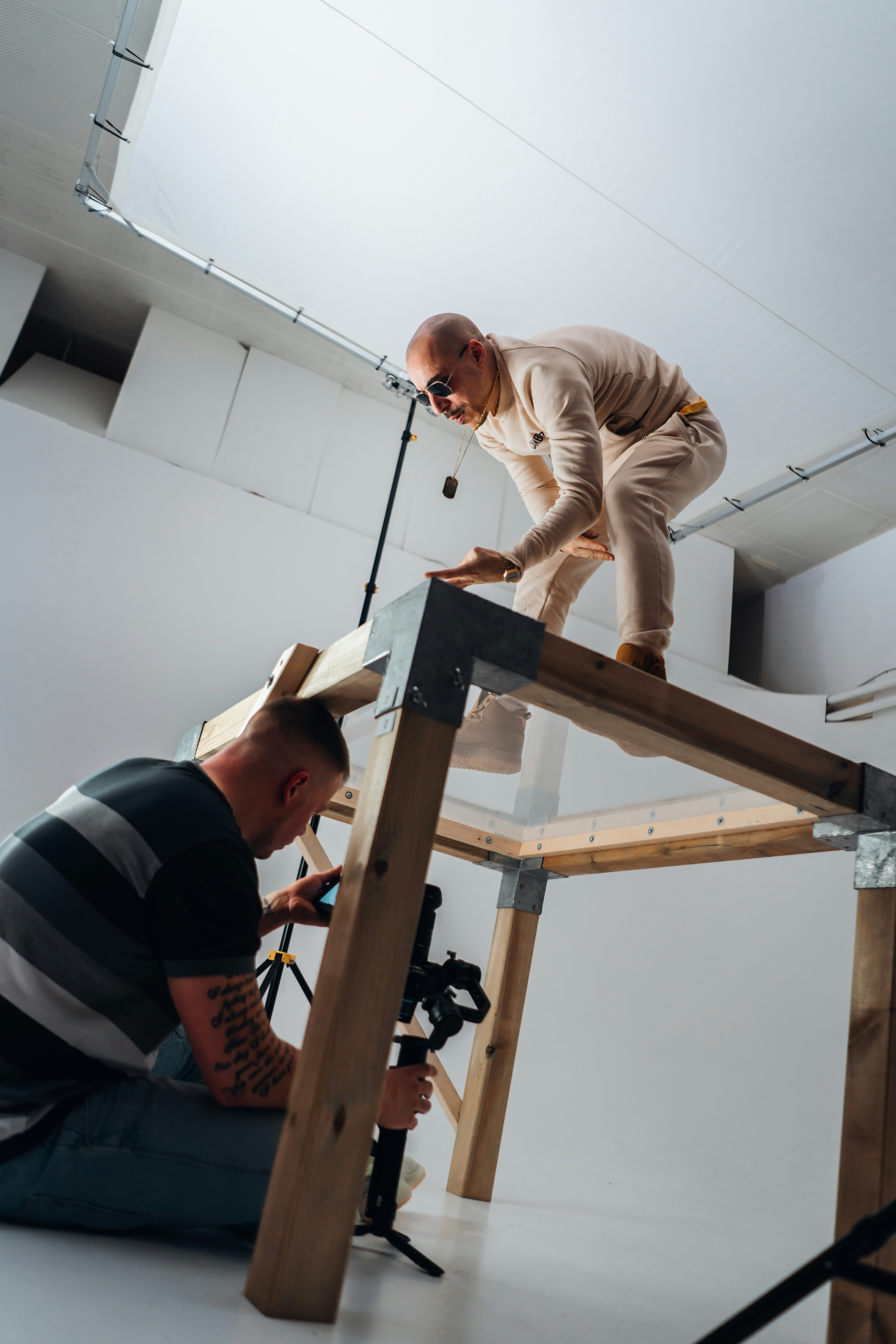 A model poses on a wooden structure while a photographer adjusts his camera setup below, highlighting the collaborative nature of studio photography.