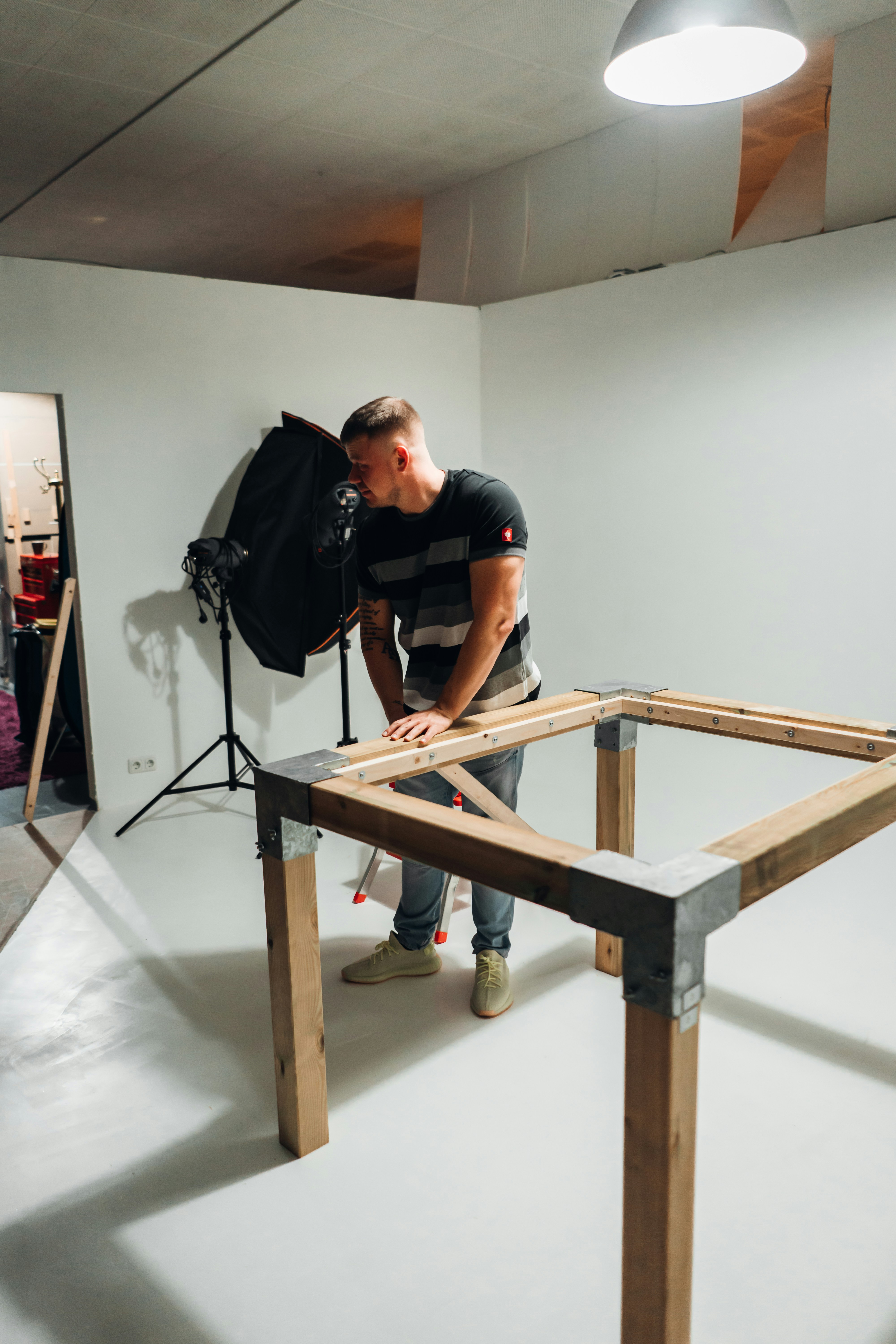 Studio scene with a man leaning over a wooden frame in a bright white room, lighting setup on a stand in the background.