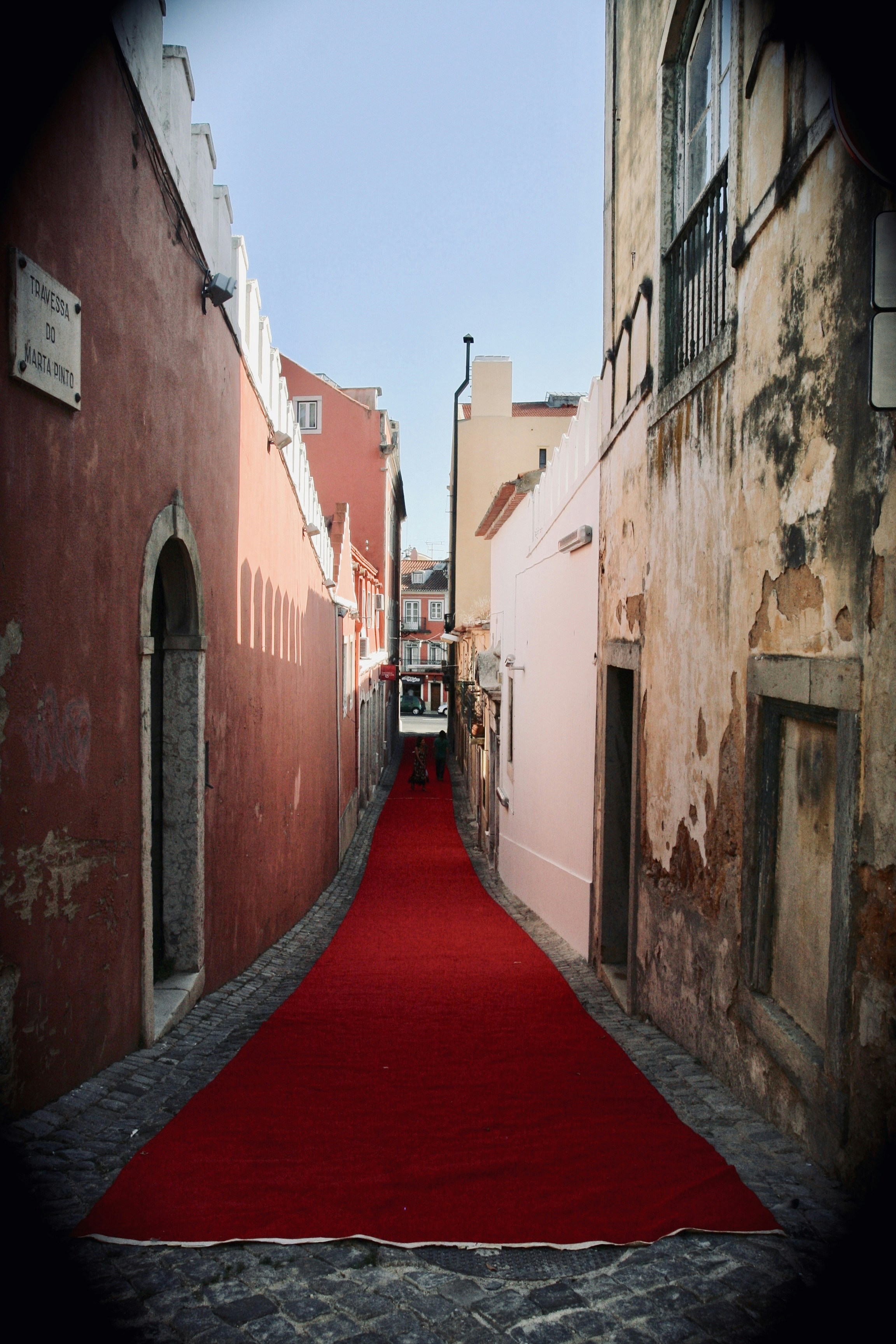 A narrow alleyway adorned with a vibrant red carpet, flanked by pastel-colored buildings under a clear blue sky.