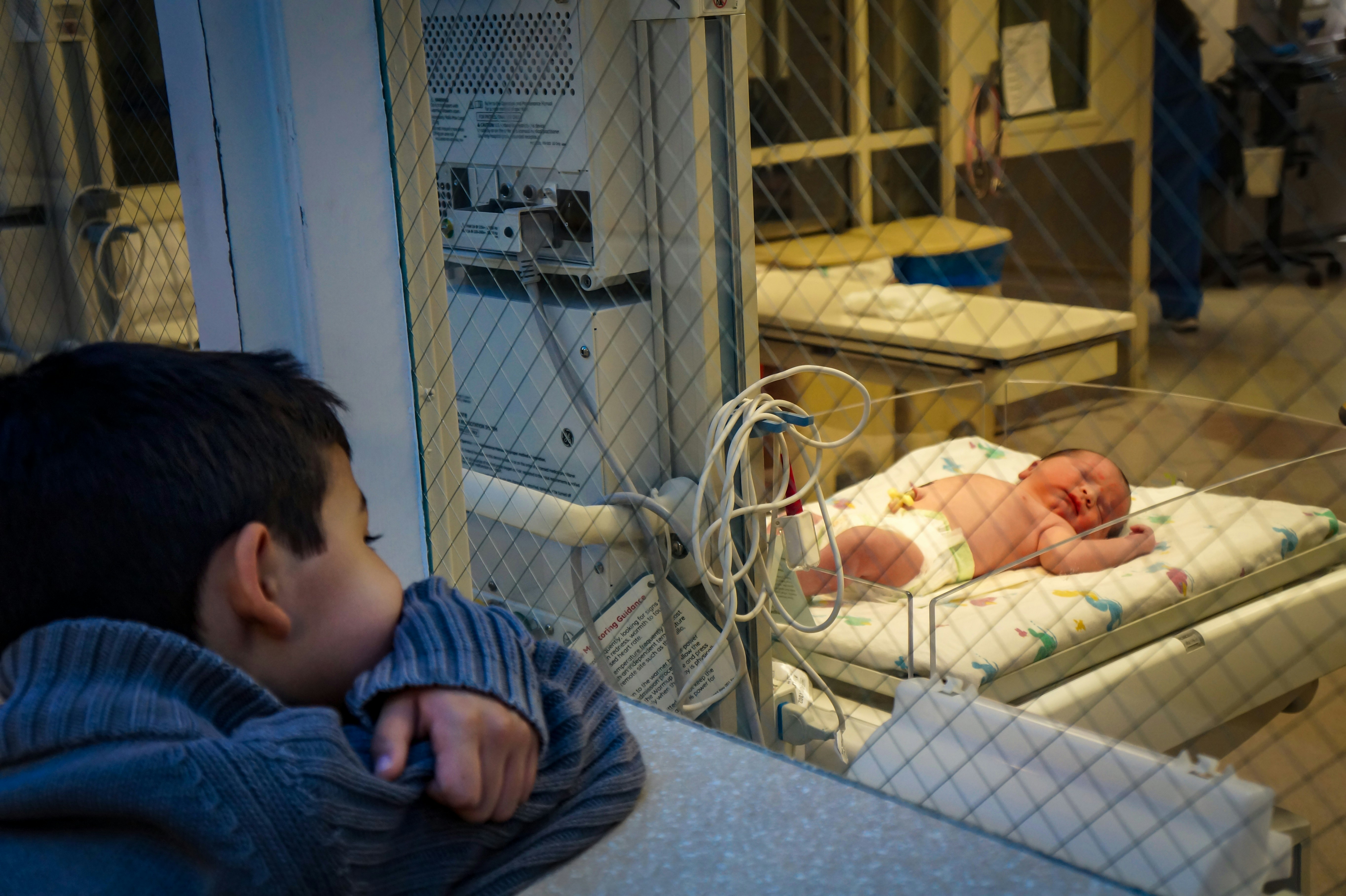 A young boy gazes through a glass partition at his newborn sibling in a hospital nursery, capturing a tender moment of familial bond.