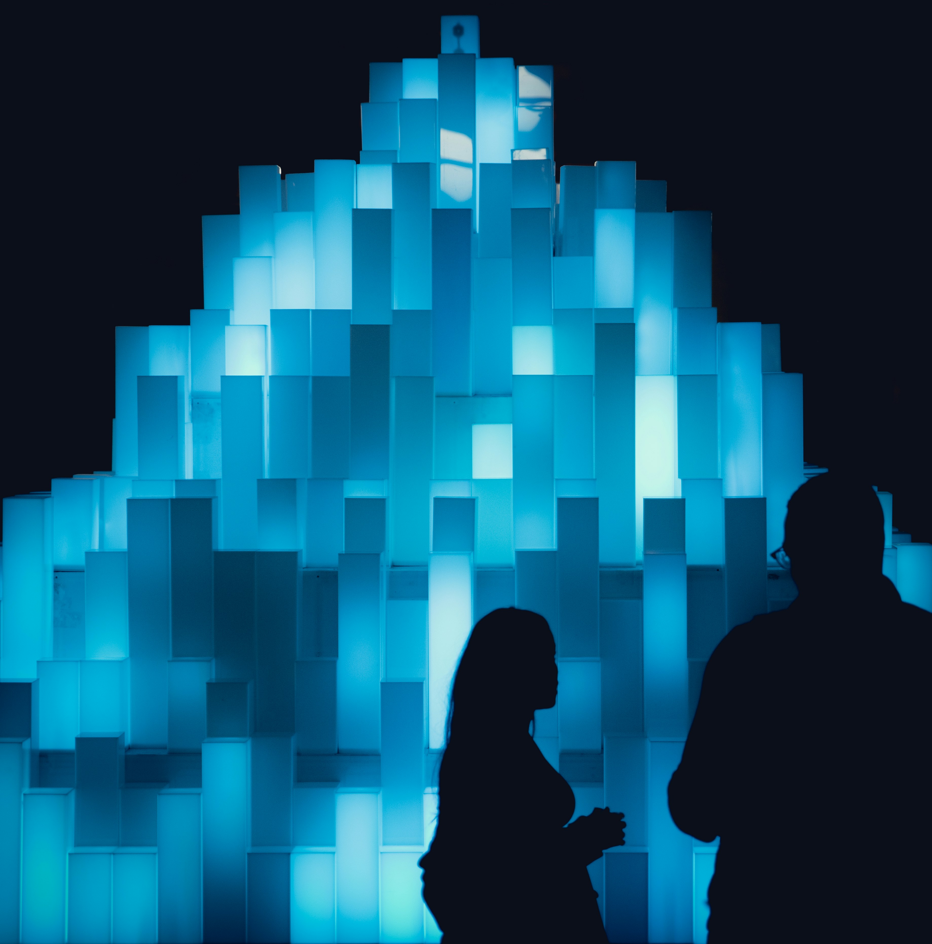 Silhouettes of two figures stand before a towering installation of illuminated blue panels, creating a striking contrast against the dark background.