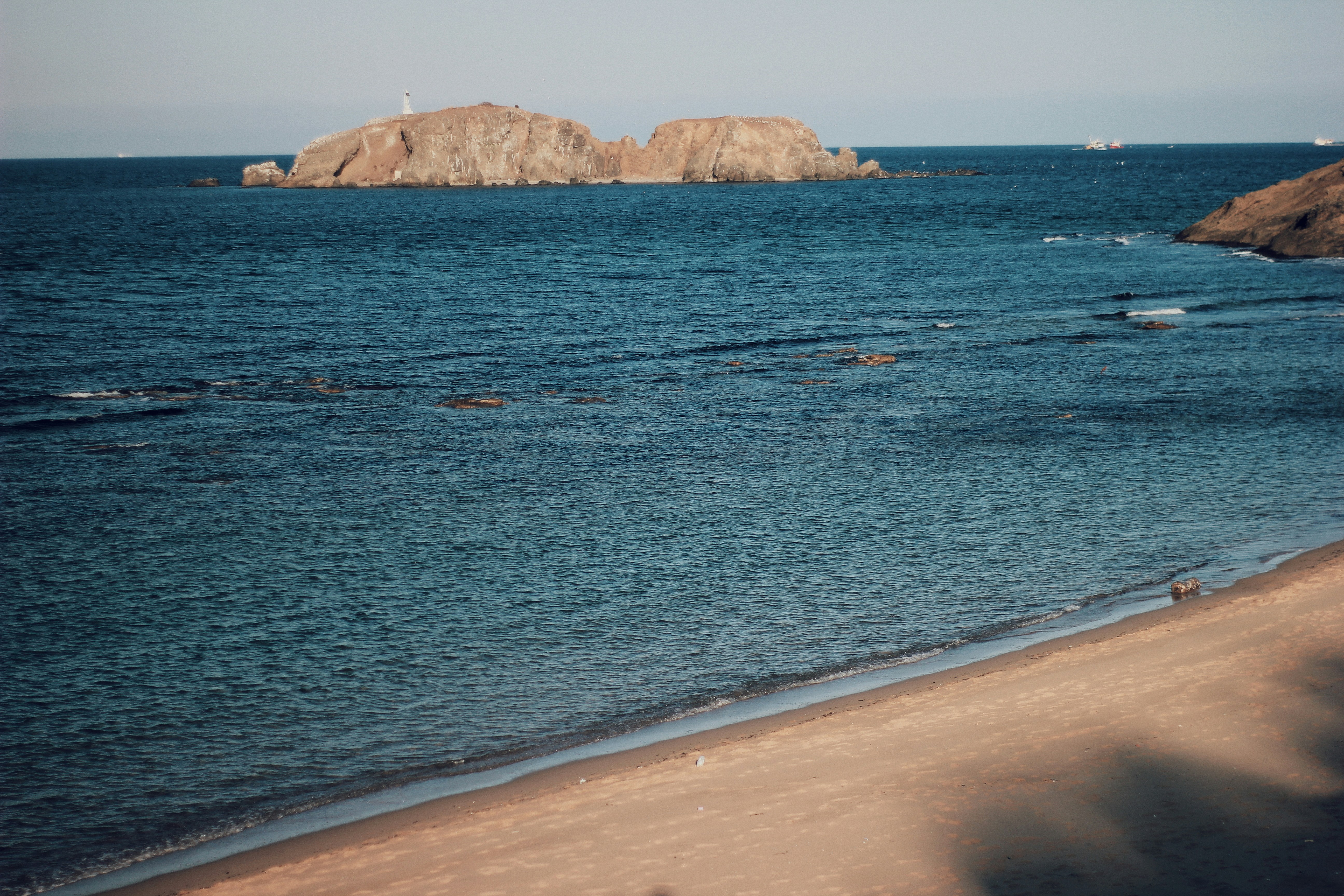 Sandy beach with gentle waves under a clear sky, leading to rocky isles on the horizon.