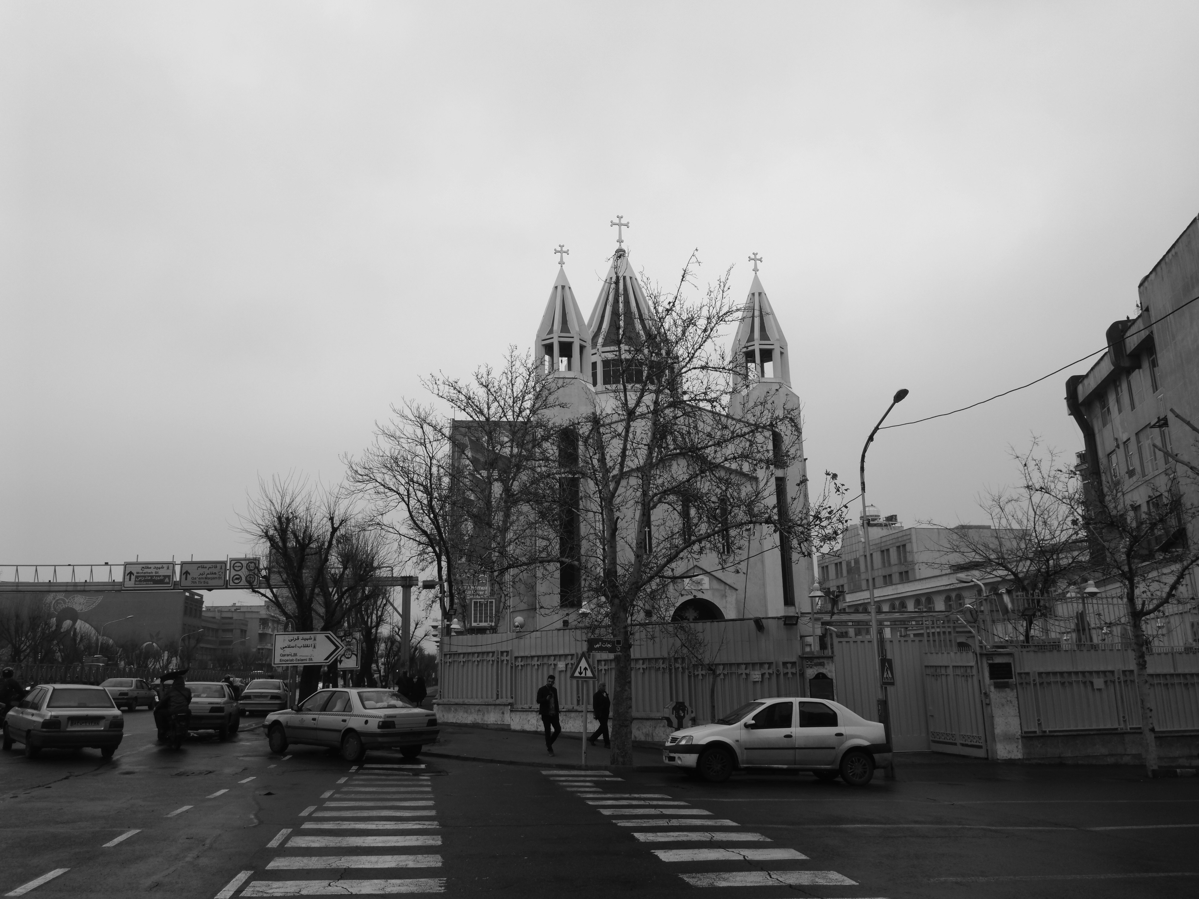 Monochrome street scene featuring a twin-spired cathedral rising behind a fence, with cars and pedestrians in the foreground.