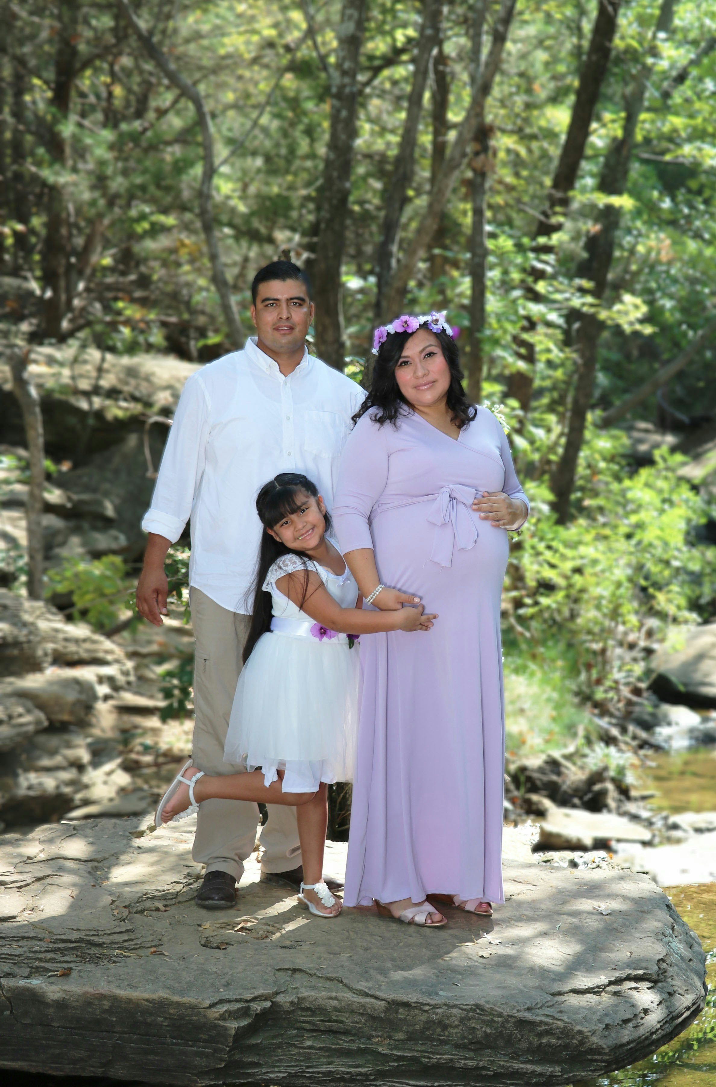 Family portrait featuring a pregnant woman in a lavender dress, a man in a white shirt, and a girl in a white dress, set against a lush, natural backdrop.