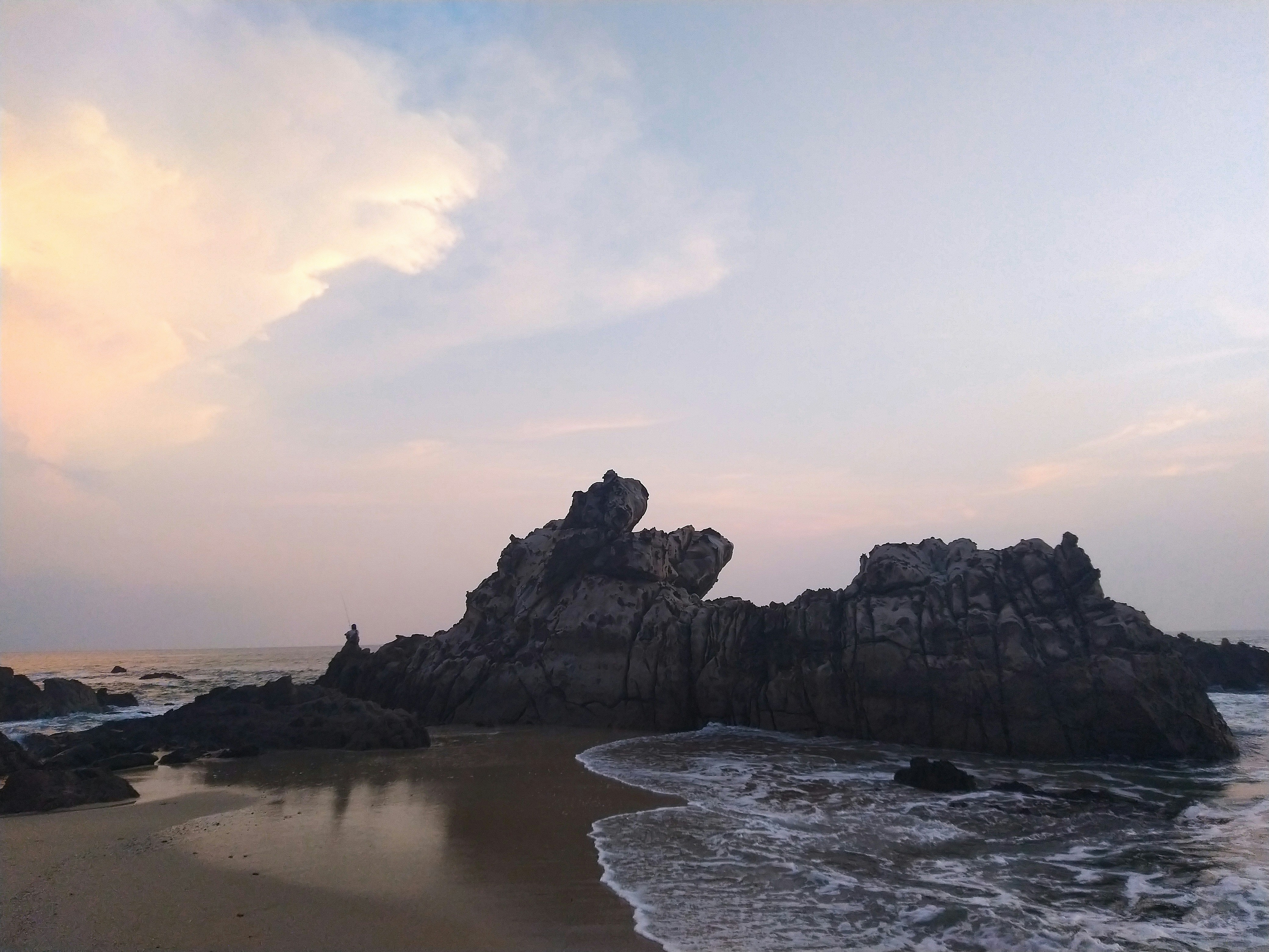 Prominent jagged rock formation rises from wet sand as gentle waves lap the shore under a pastel sunset.