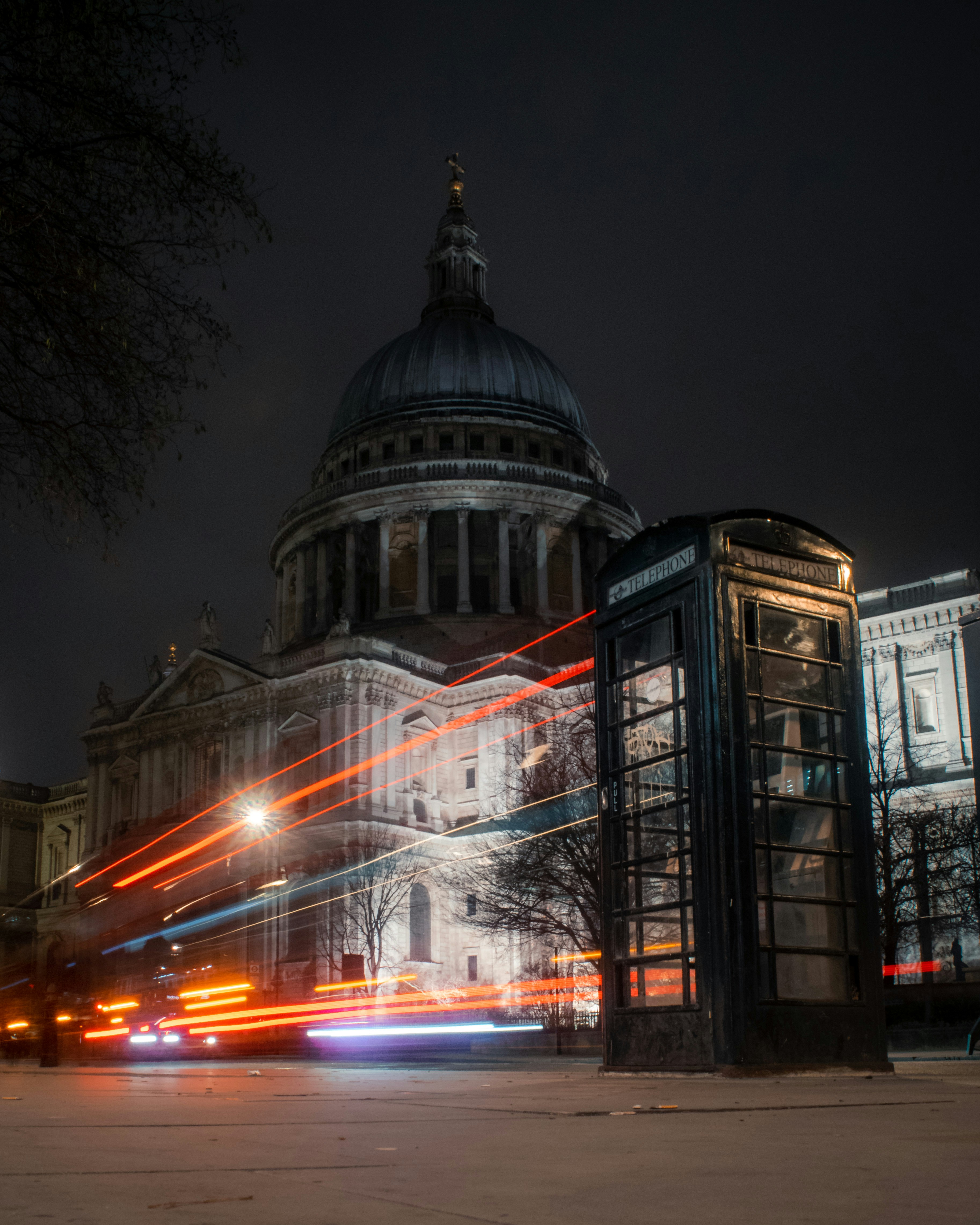 Historic telephone booth stands in front of St. Paul's Cathedral, illuminated by streaks of passing vehicle lights at night.