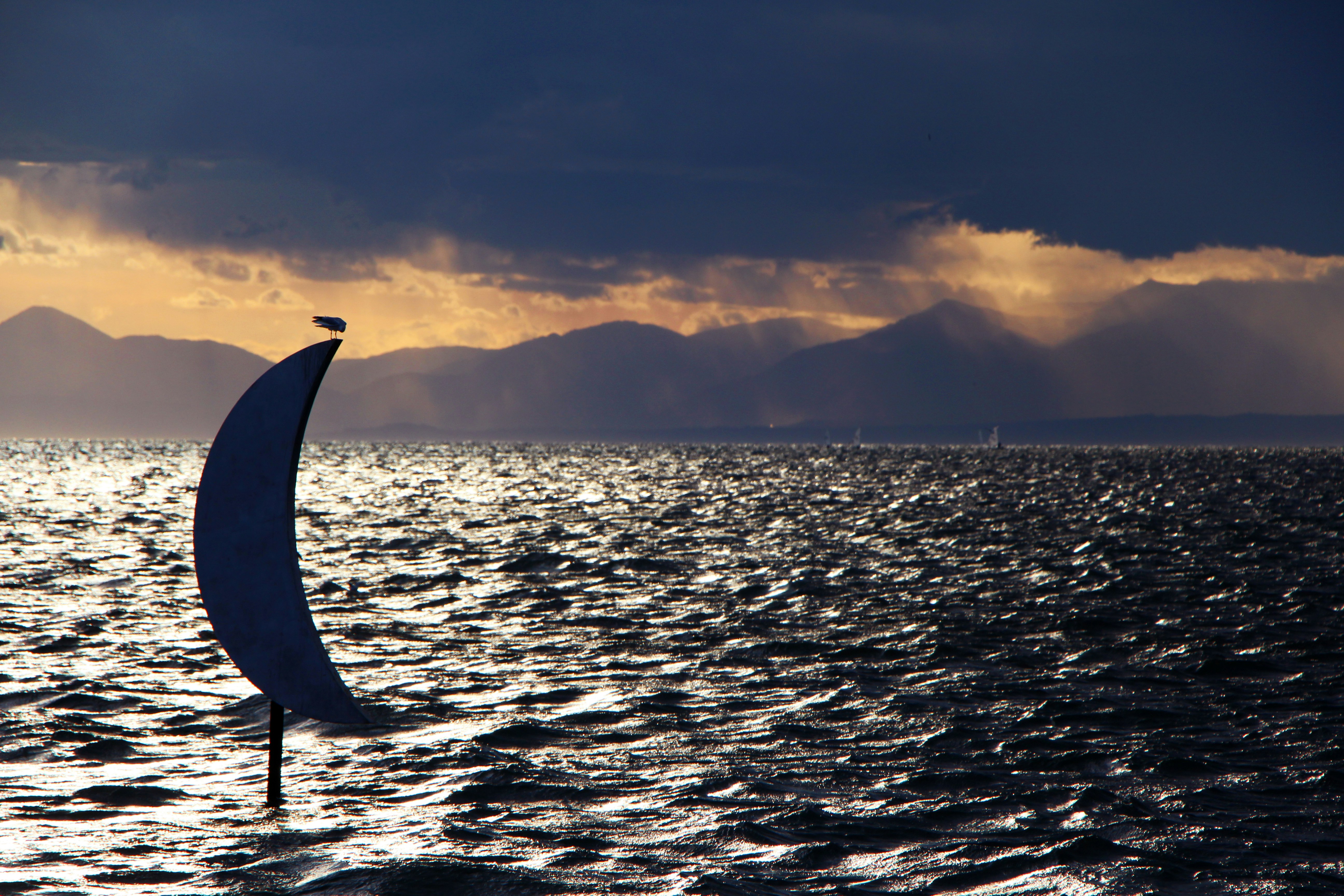 Sailboat with a curved sail gliding through shimmering water under a dramatic dusk sky.