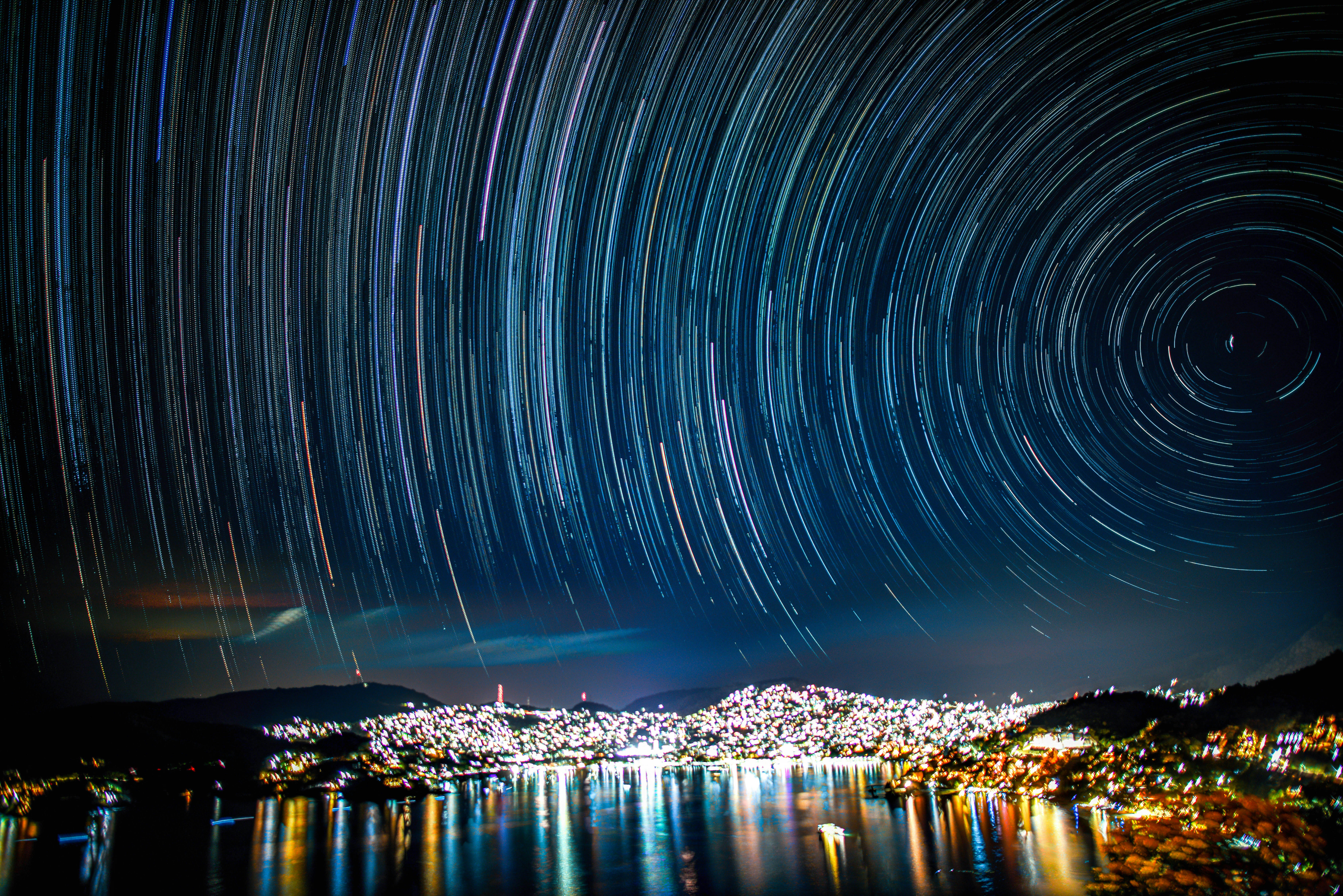 Star trails create circular patterns over a brightly lit coastal town at night.