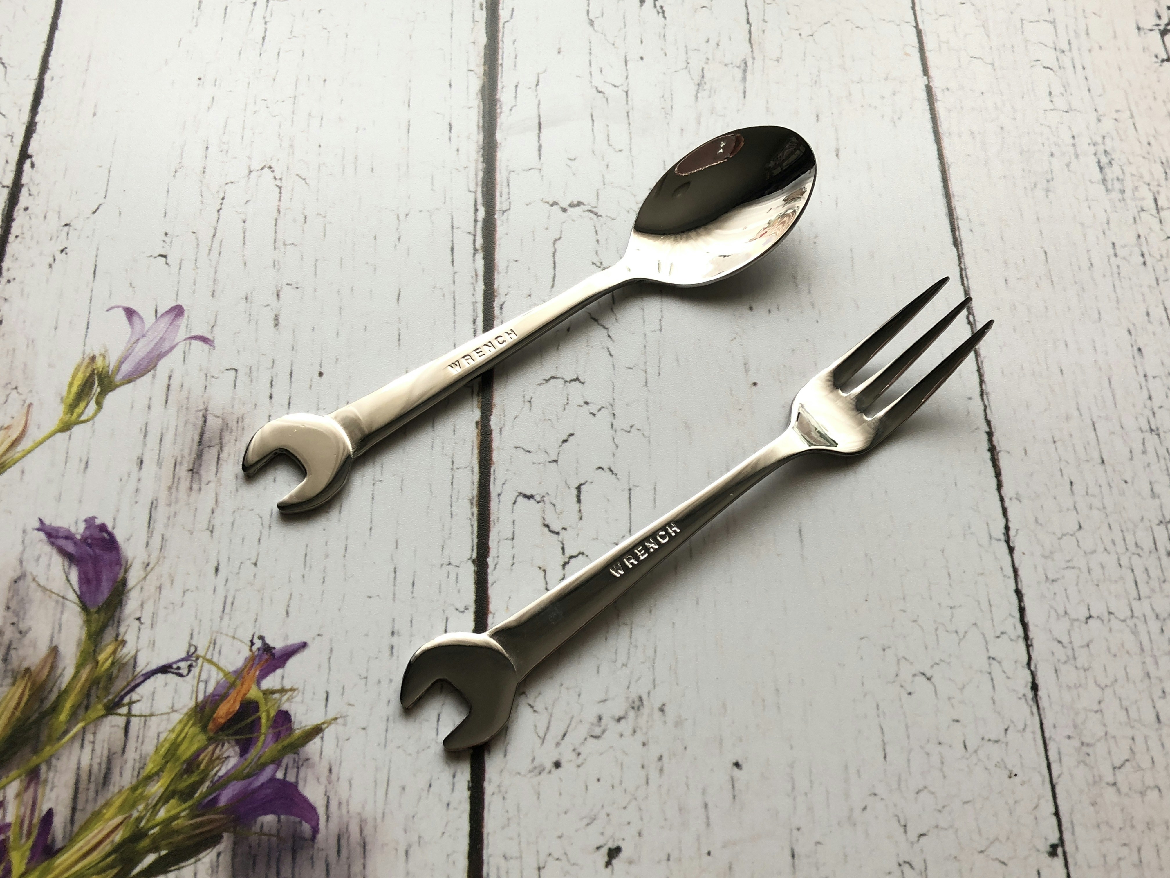 Unique fork and spoon designed to resemble wrenches, resting on a rustic wooden surface with floral accents.