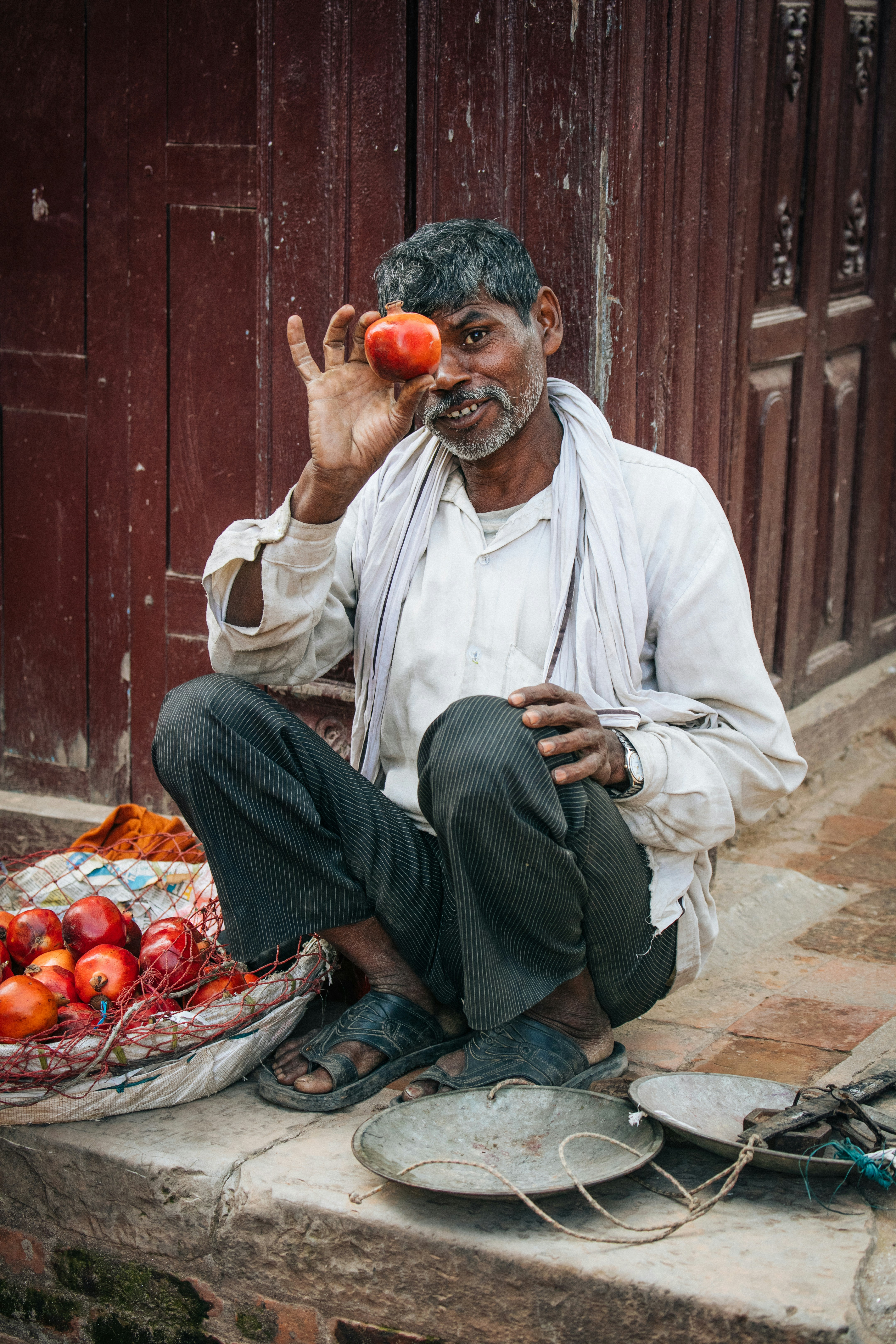 Elderly vendor showcasing a ripe tomato while seated beside a basket of fresh produce against a textured wooden backdrop.