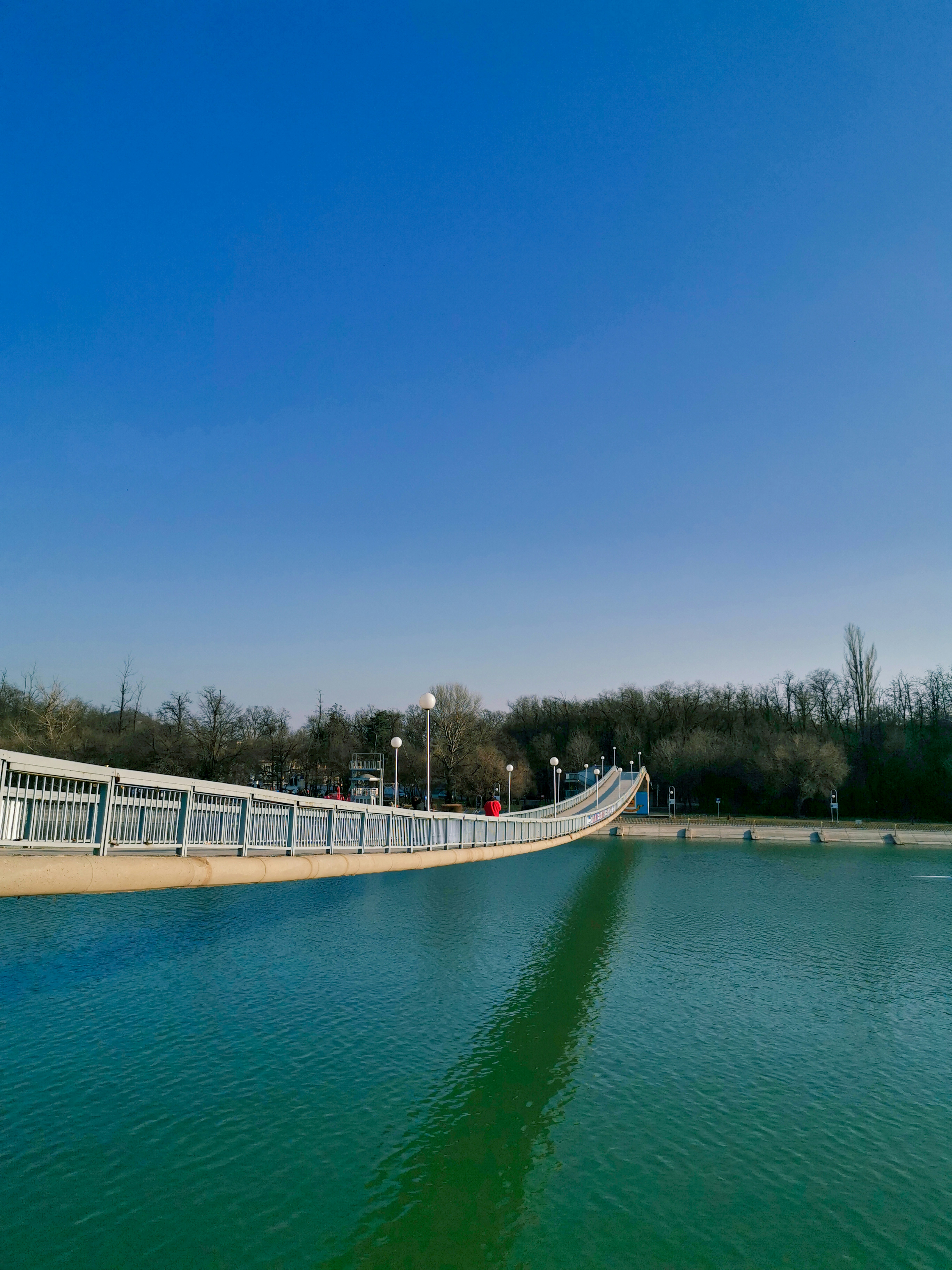 Curved pedestrian bridge spanning a tranquil body of water under a clear blue sky.