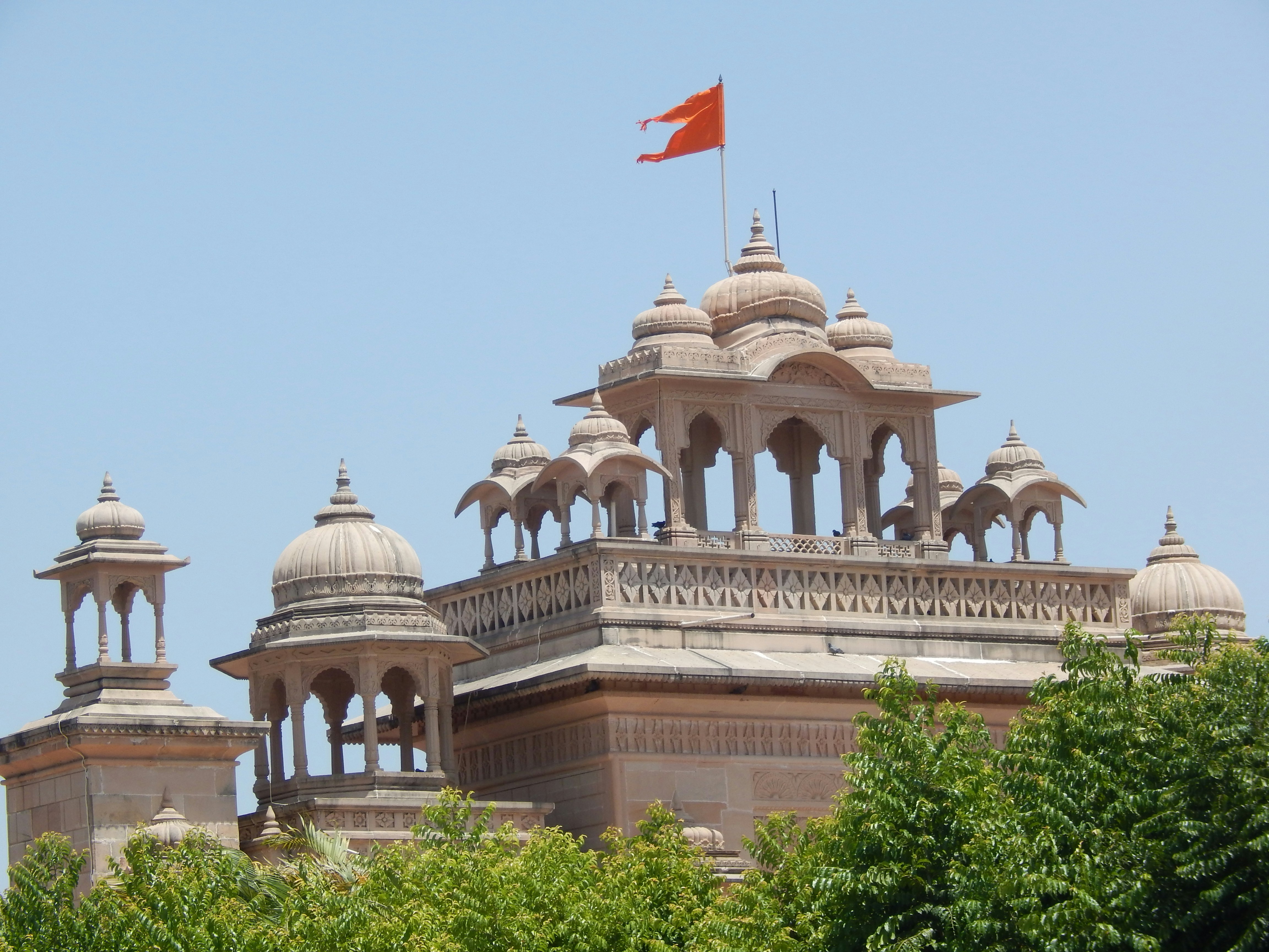 Ornate temple domes with a vibrant flag atop, surrounded by lush greenery against a clear blue sky.