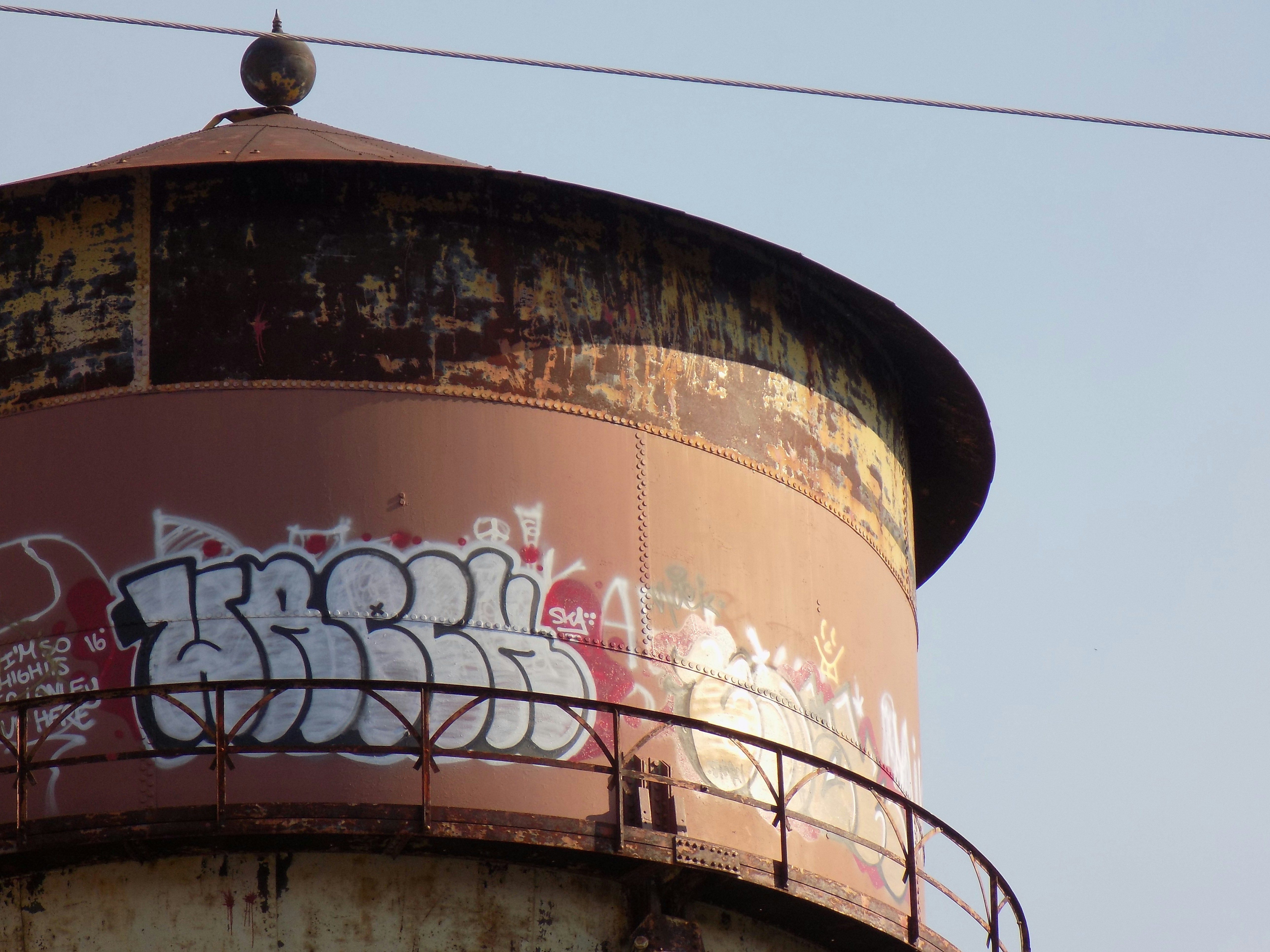 An aged rust-colored cylindrical tank is wrapped in vivid graffiti against a pale blue sky. A metal railing circles its midsection as power lines stretch across the frame.