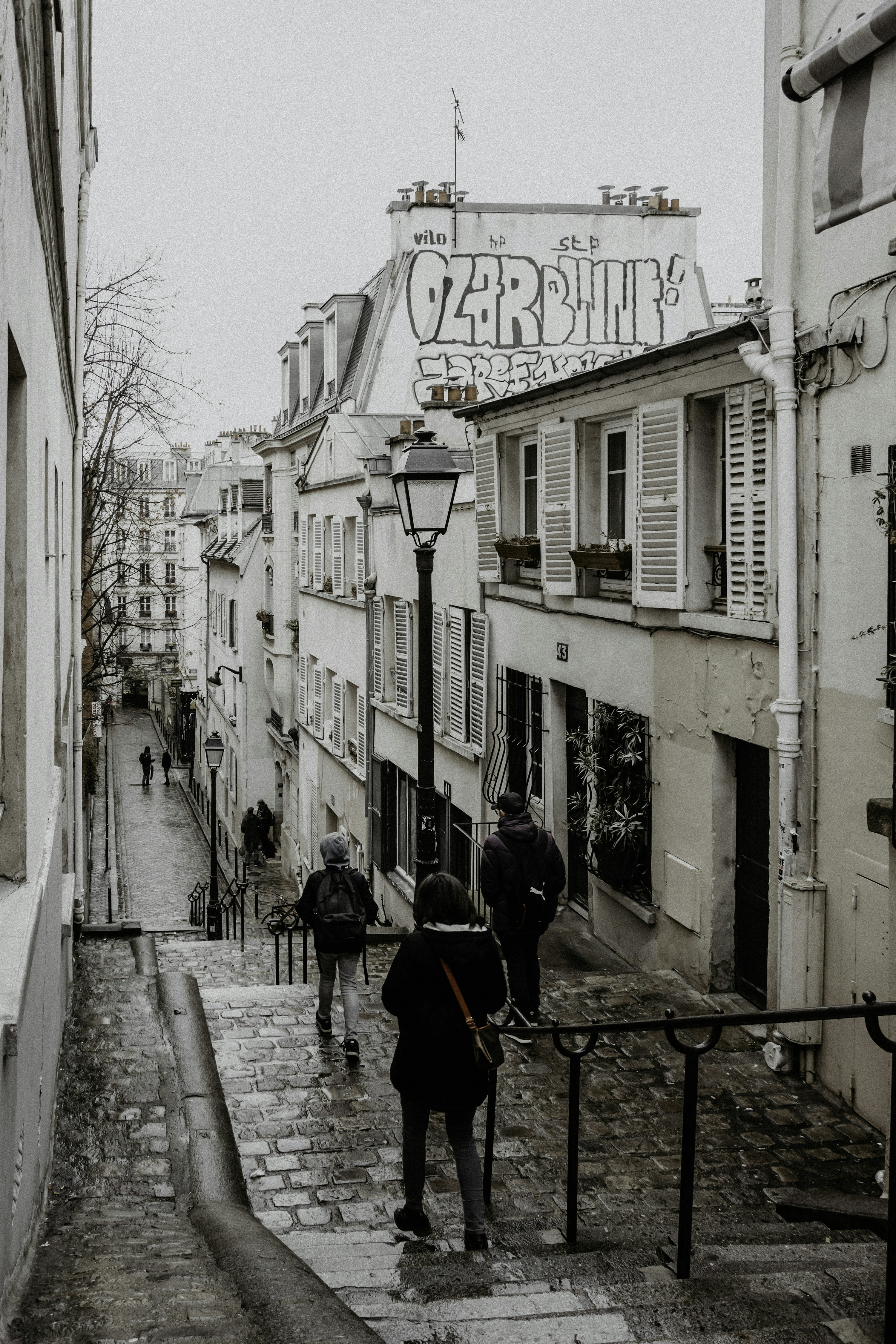 Cobblestone street in Montmartre, Paris, with people walking under a cloudy sky and street lamps illuminating the path.