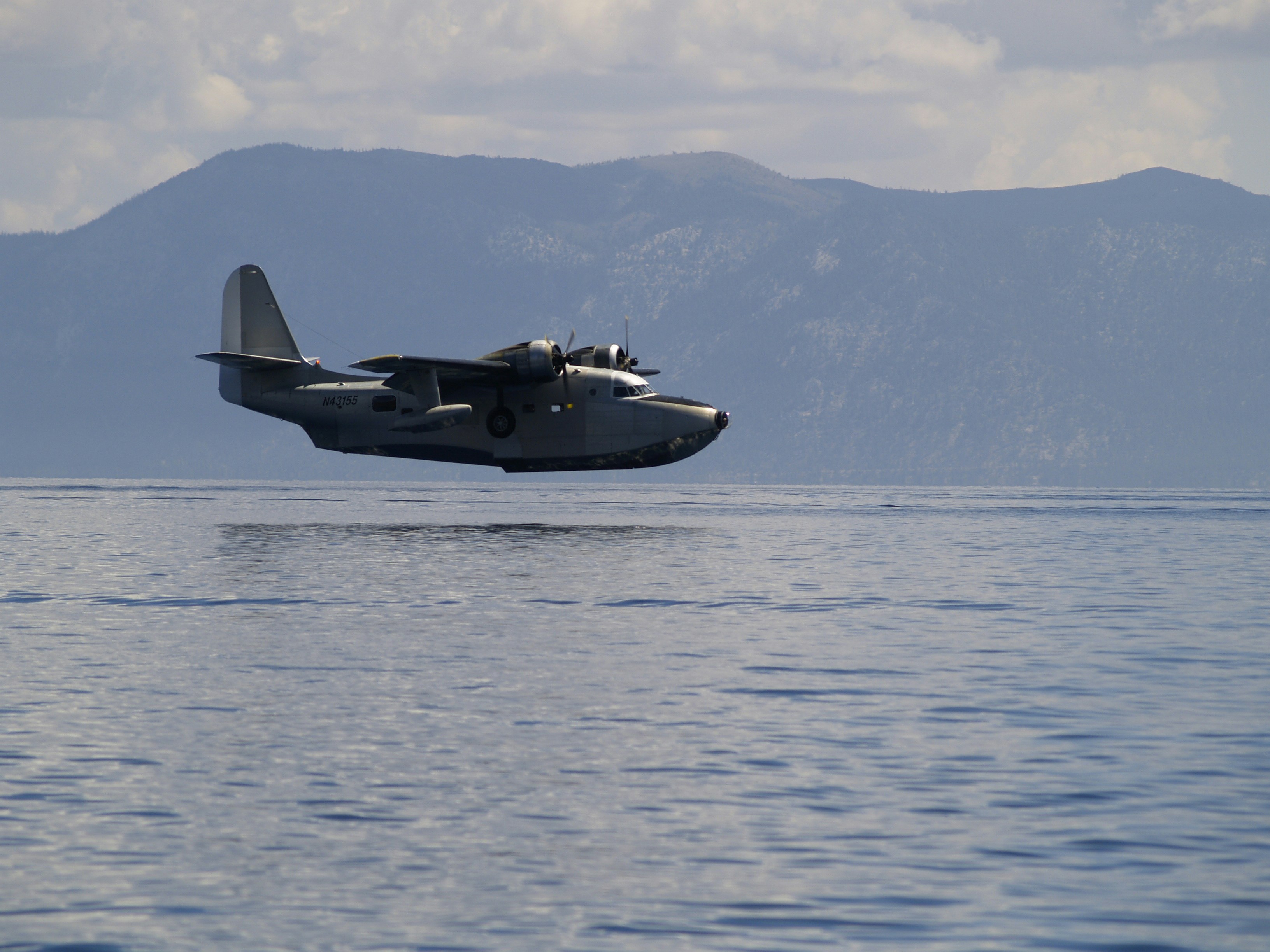 Amphibious aircraft gliding gracefully above a tranquil lake, framed by distant mountains under a partly cloudy sky.