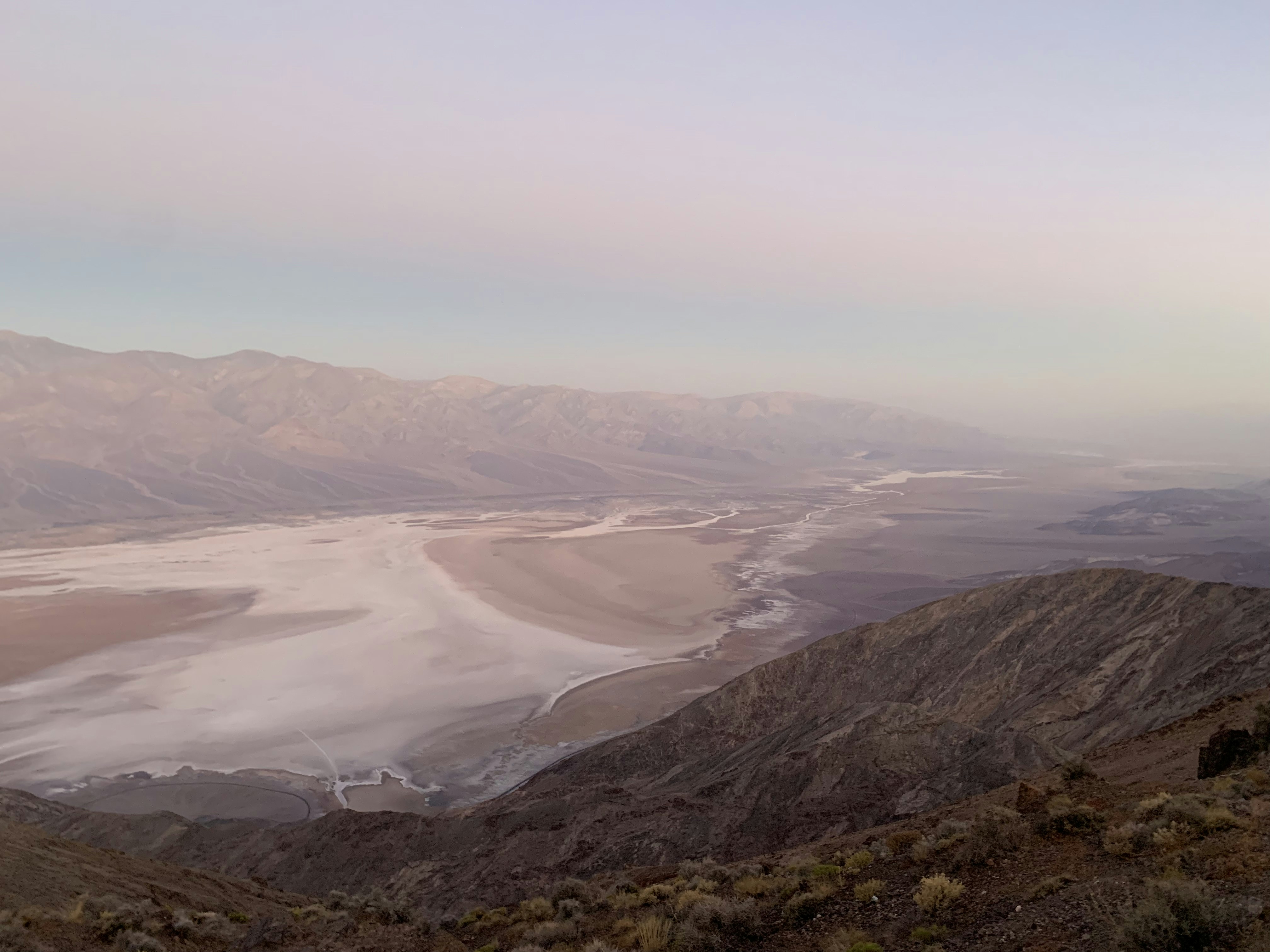 Expansive view of a desert valley with a winding river and salt flats under a pastel sky at dusk.