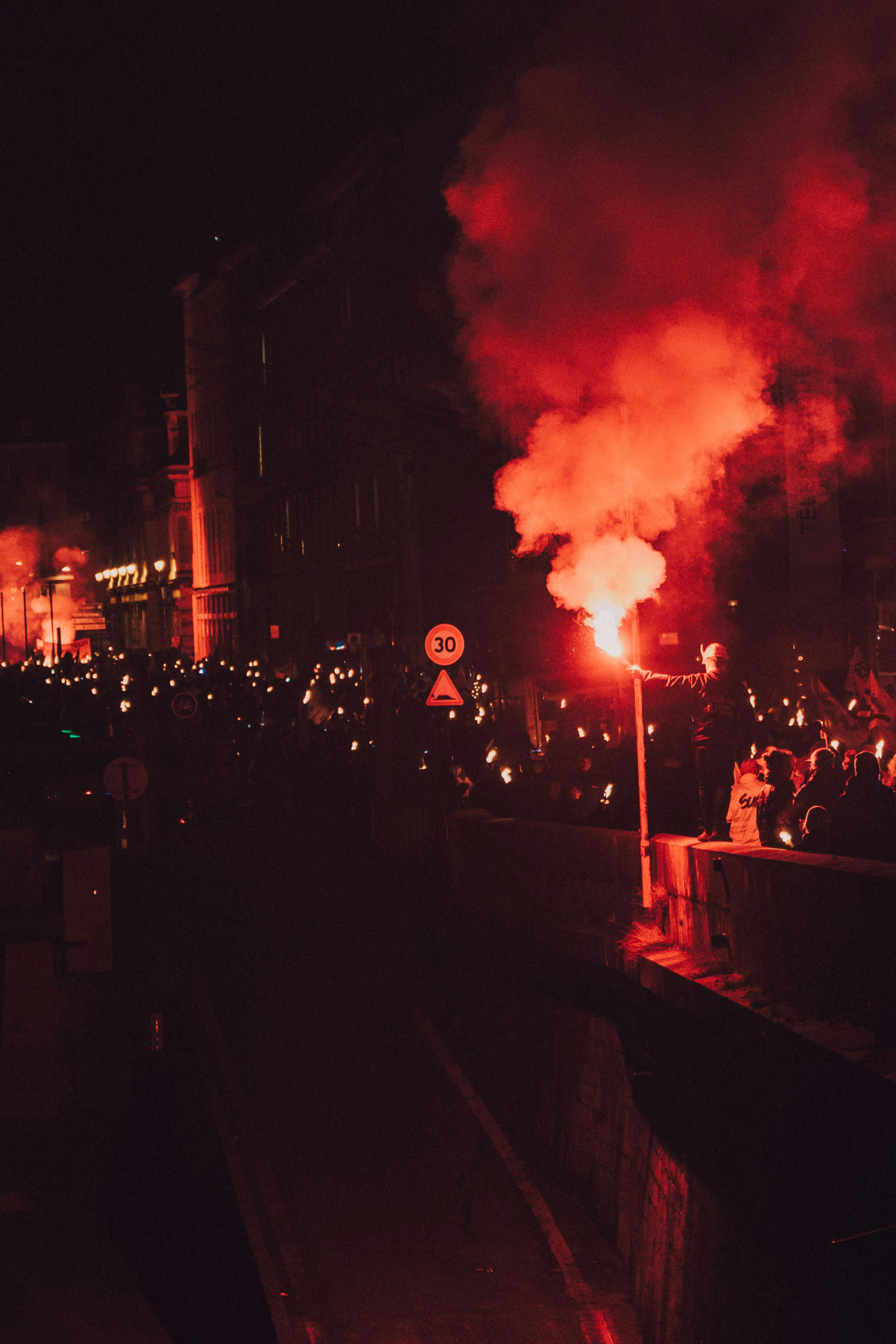 Crowd holding flares during a nighttime celebration, illuminated by vibrant red smoke and lights. The scene captures the energy of a festive gathering.