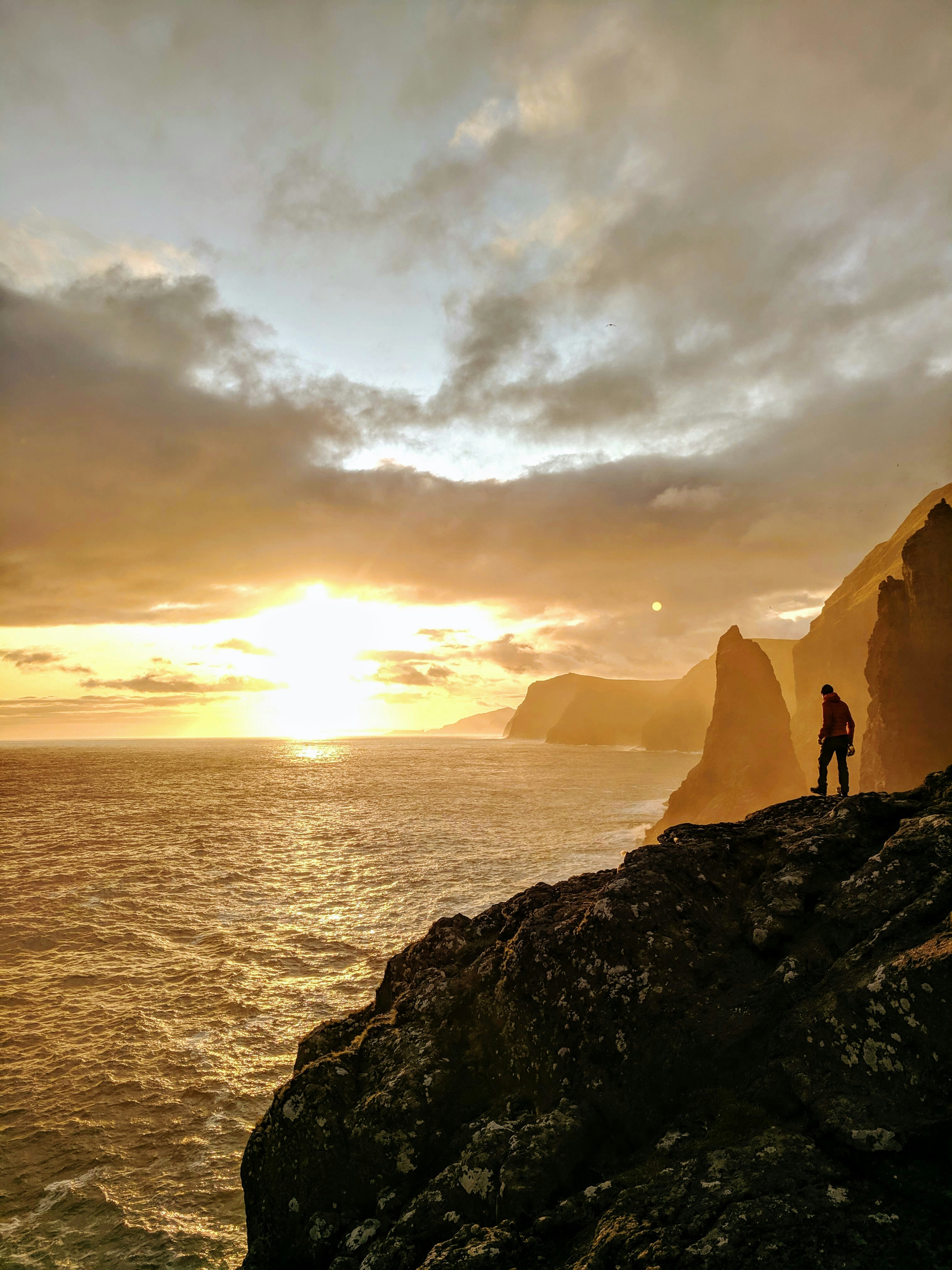 A lone figure stands atop a rugged cliff, gazing at the sunset over the ocean, with dramatic rock formations framing the scene.