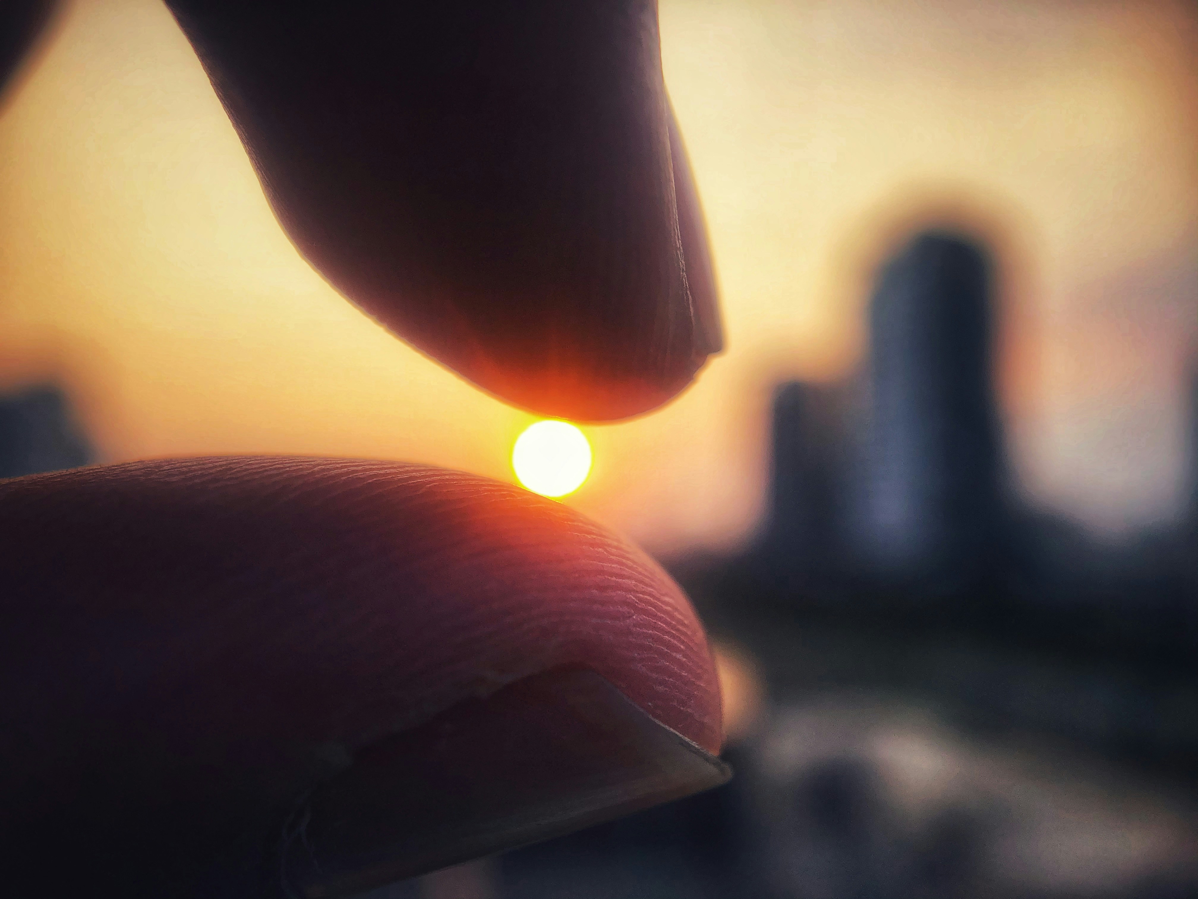 Fingers gently cradle the sun against a backdrop of blurred skyscrapers at sunset.