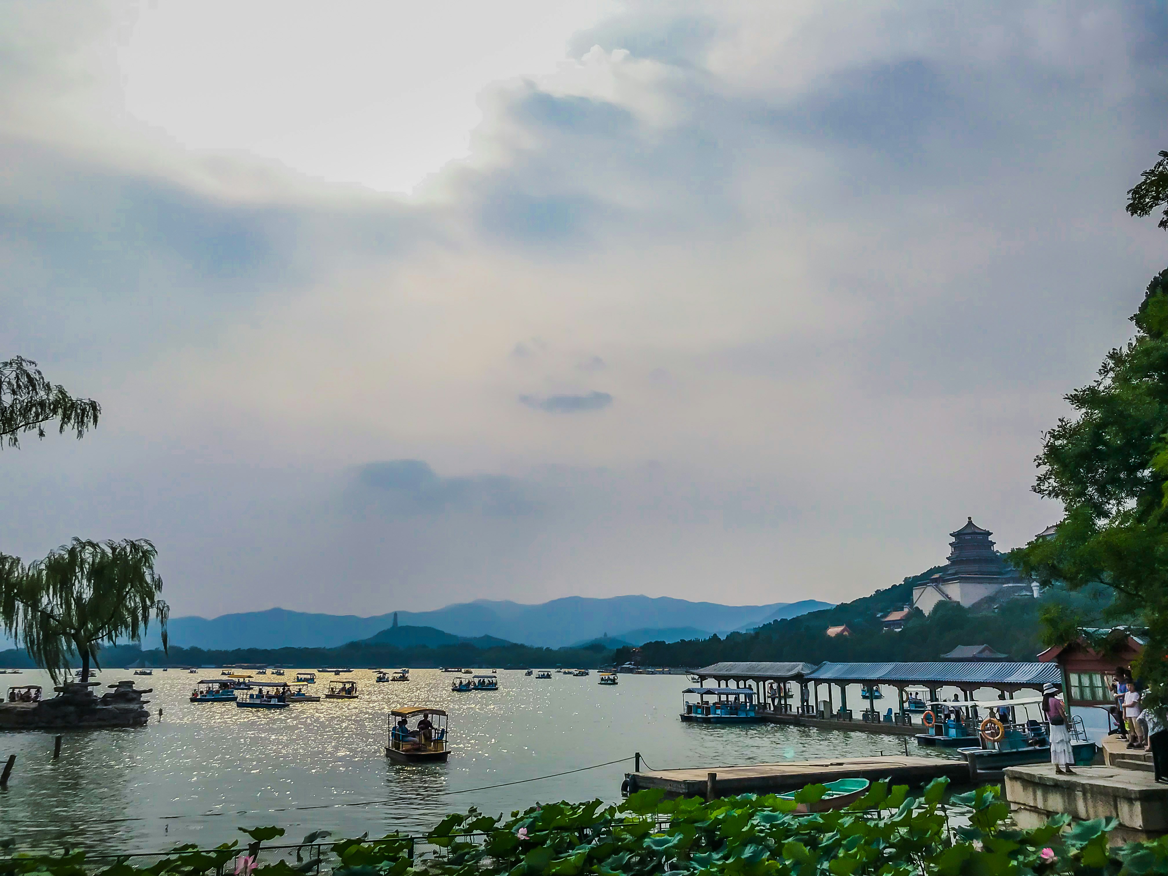 Boats gently gliding on a tranquil lake, framed by distant mountains and a historic pavilion under a cloudy sky.