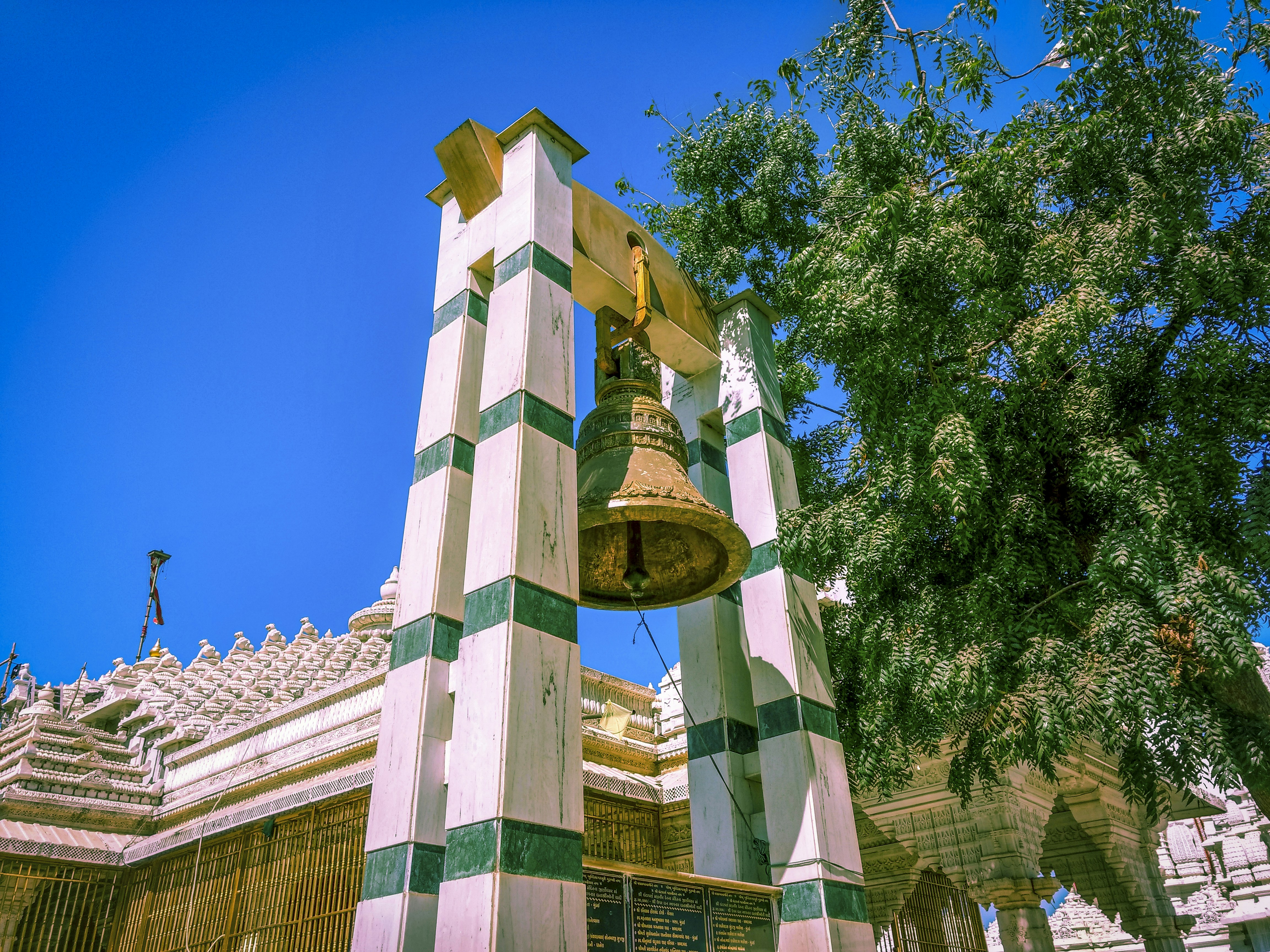 A large bell hangs from a strikingly designed structure adorned with green and white stripes, set against a clear blue sky near a temple complex.
