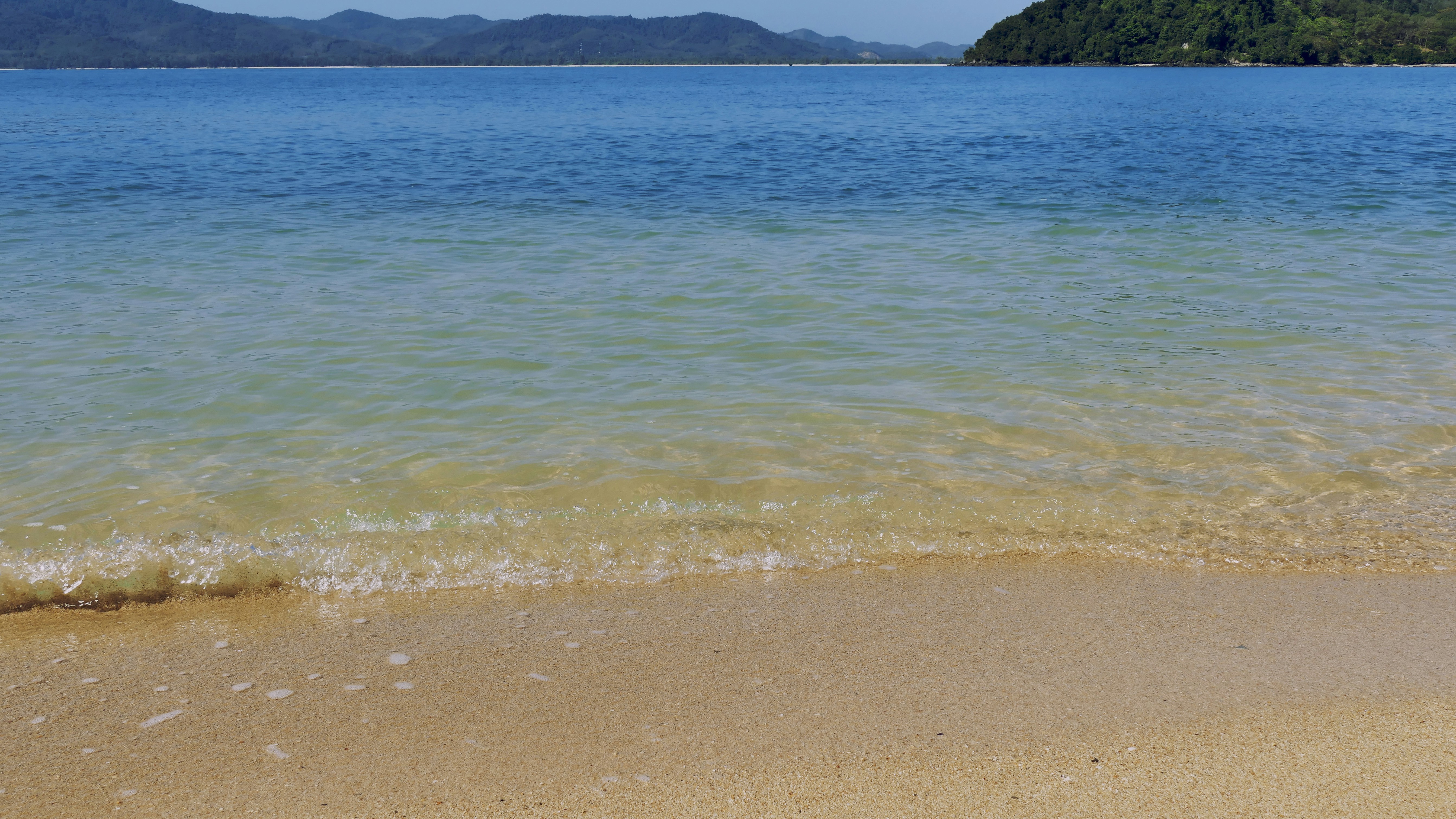 Gentle waves lapping against a sandy beach under a clear blue sky, with distant green hills on the horizon.
